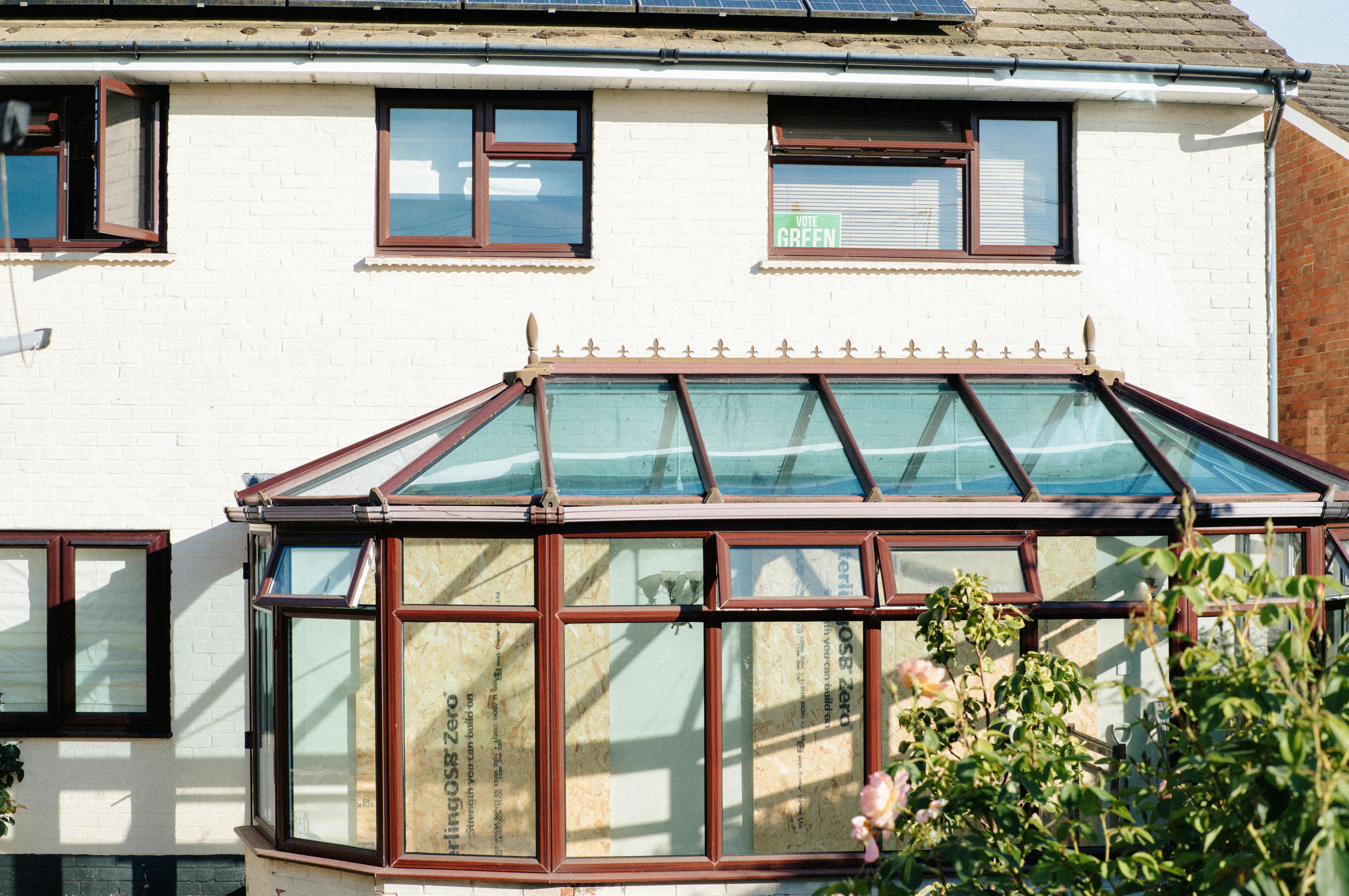 Bright white brick house with a large glass conservatory spanning the lower half and red-brown window frames; greenery in the foreground hints at a garden setting.