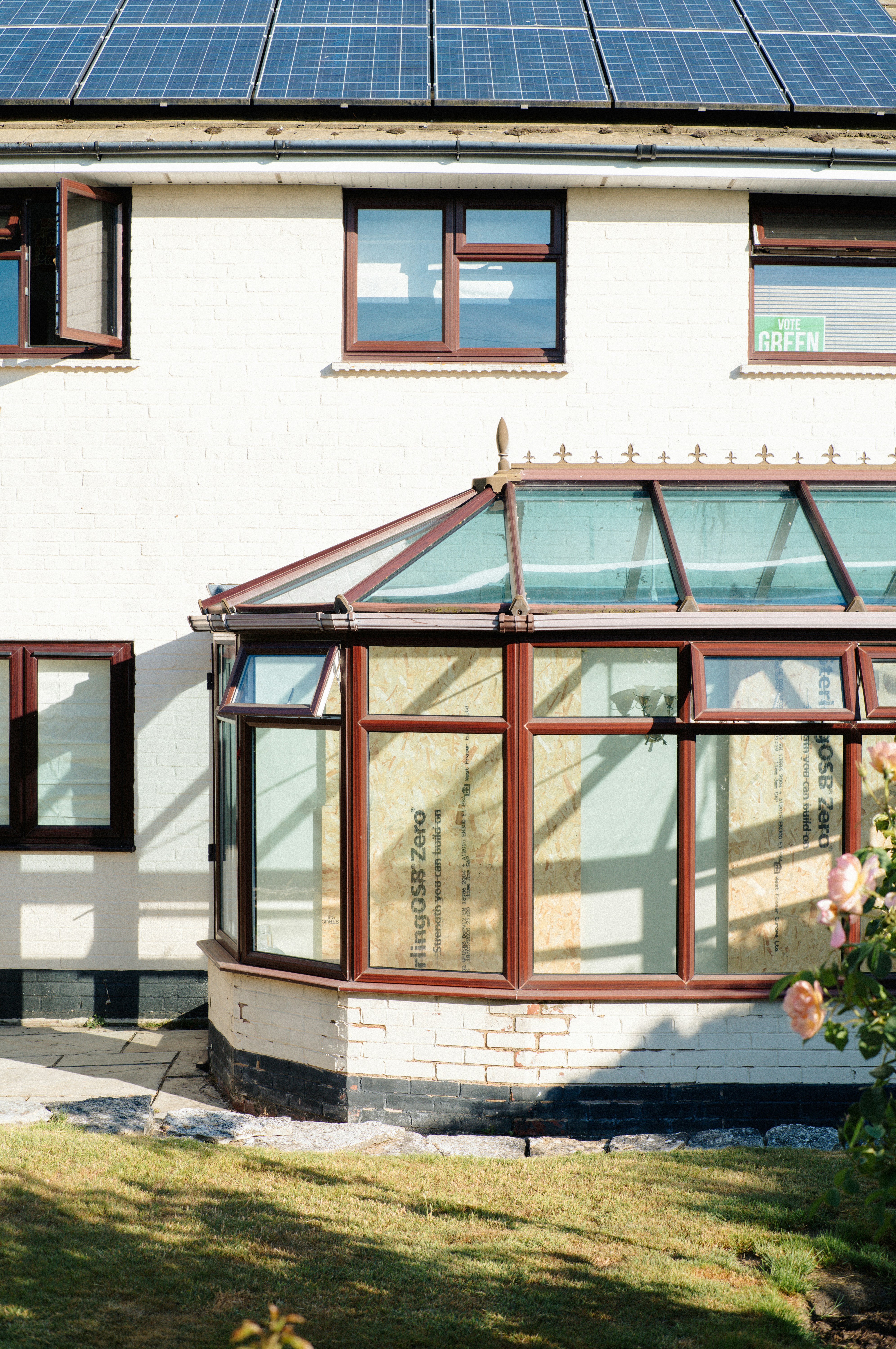 Bright white house with a glass conservatory and solar panels on the roof; the scene centers on the sunlit conservatory bays and their wooden frames.