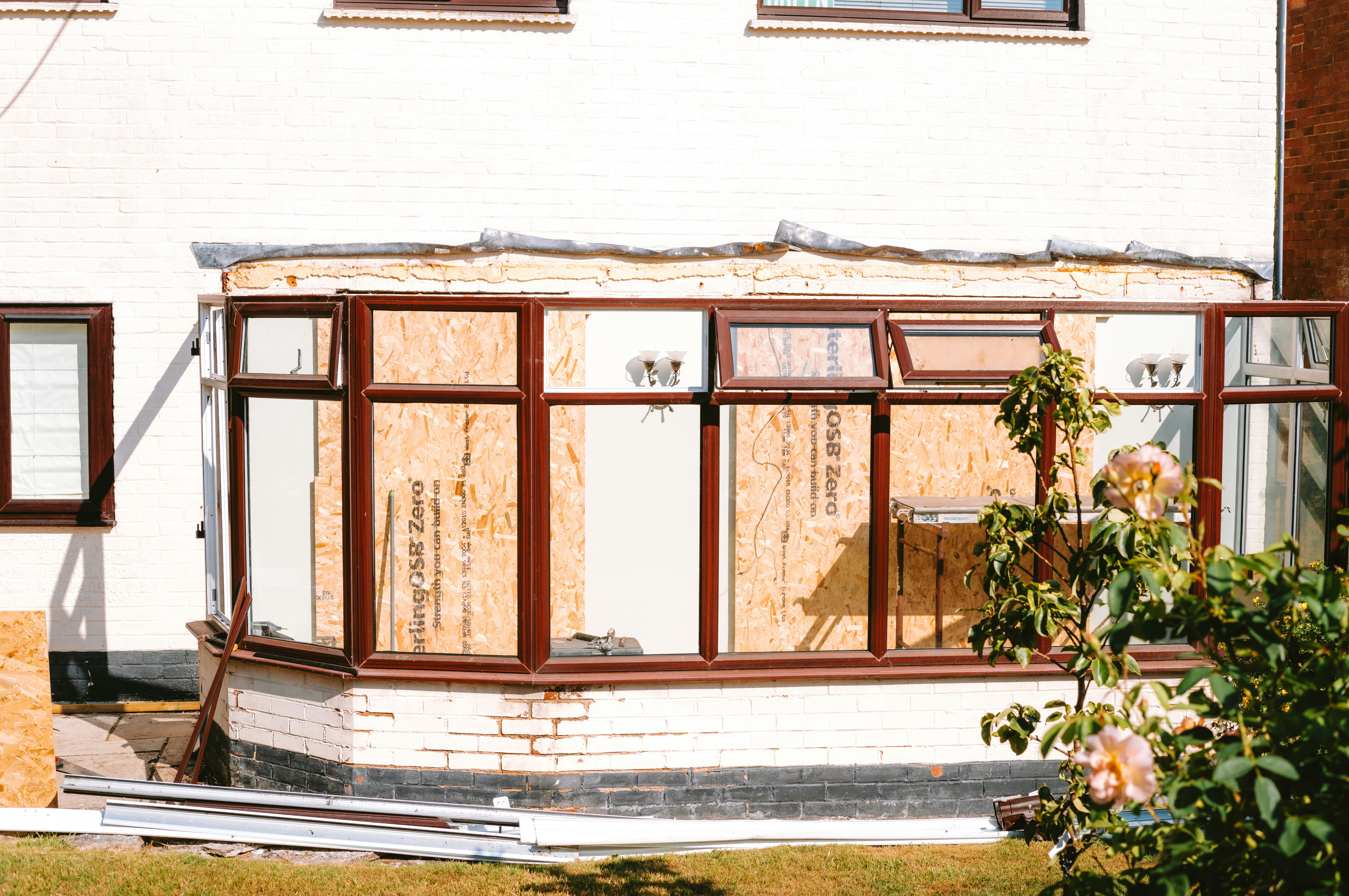 Partially boarded conservatory with exposed framing and a rose bush in the foreground; construction materials and plywood sheathing visible through the windows.