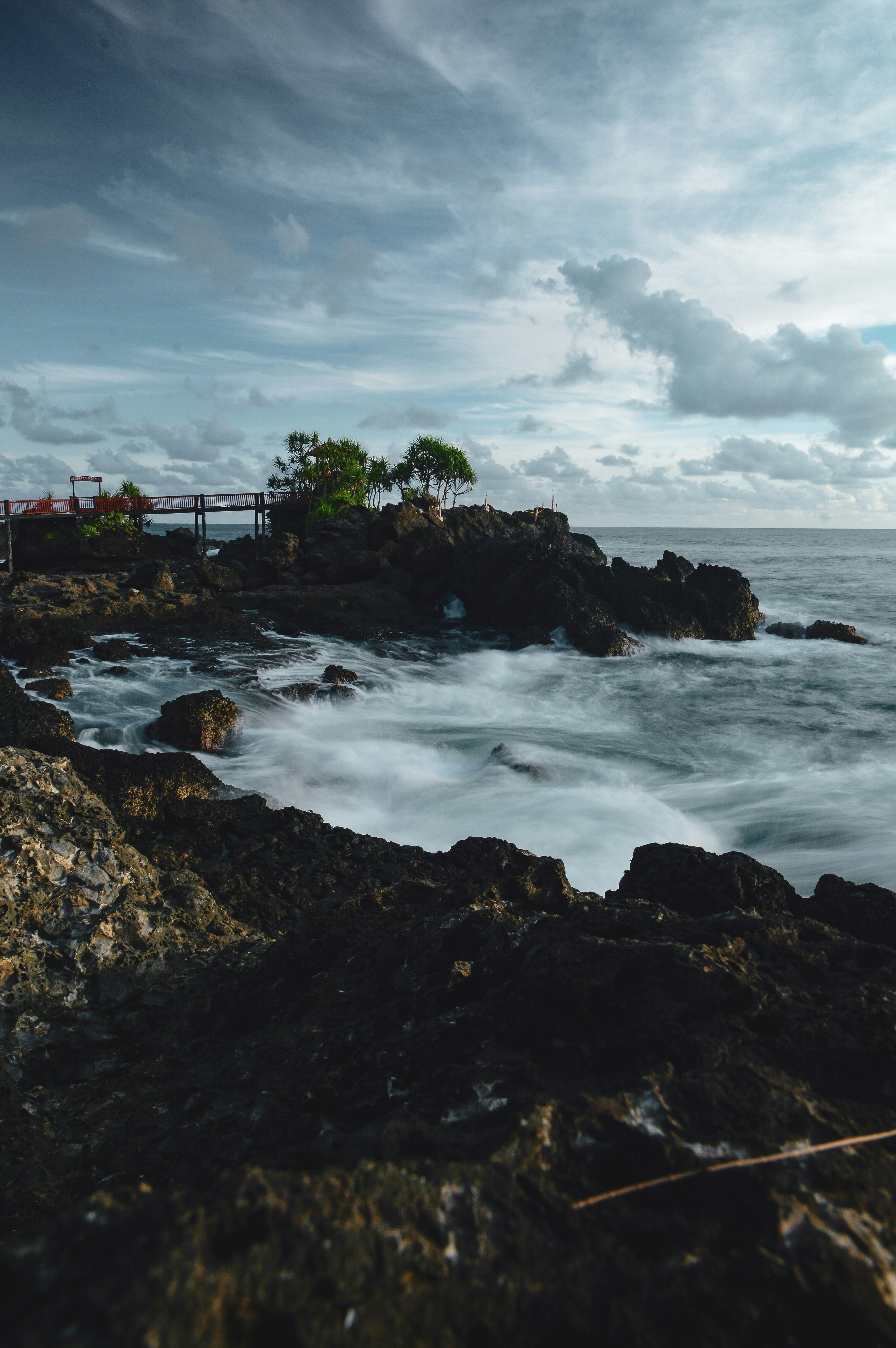 Rugged coastline with dark volcanic rocks meeting foamy sea, small lookout and trees on a rocky outcrop under a moody sky.