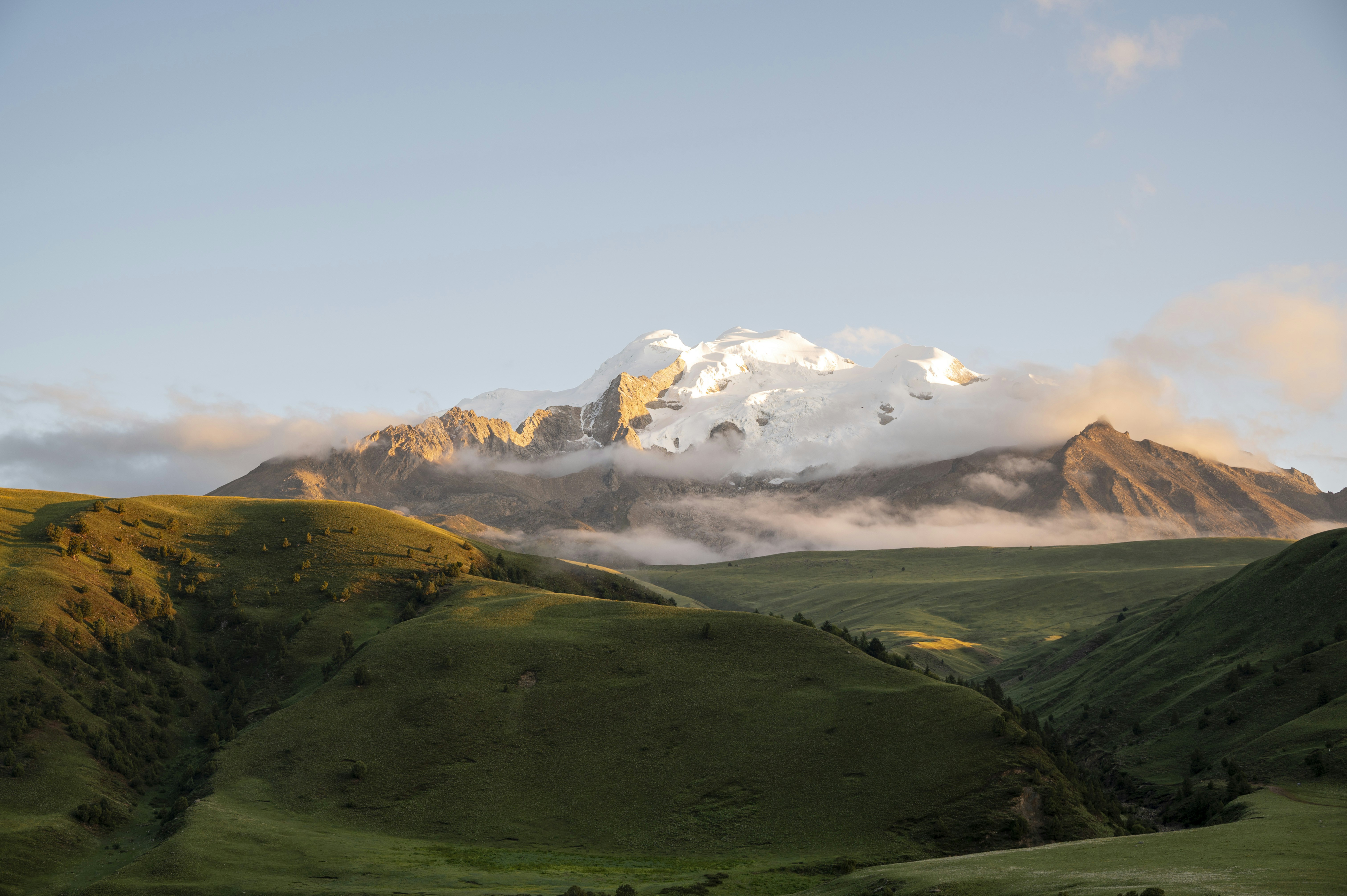 Snow-capped summit breaking through a low-lying cloud belt over rolling green foothills