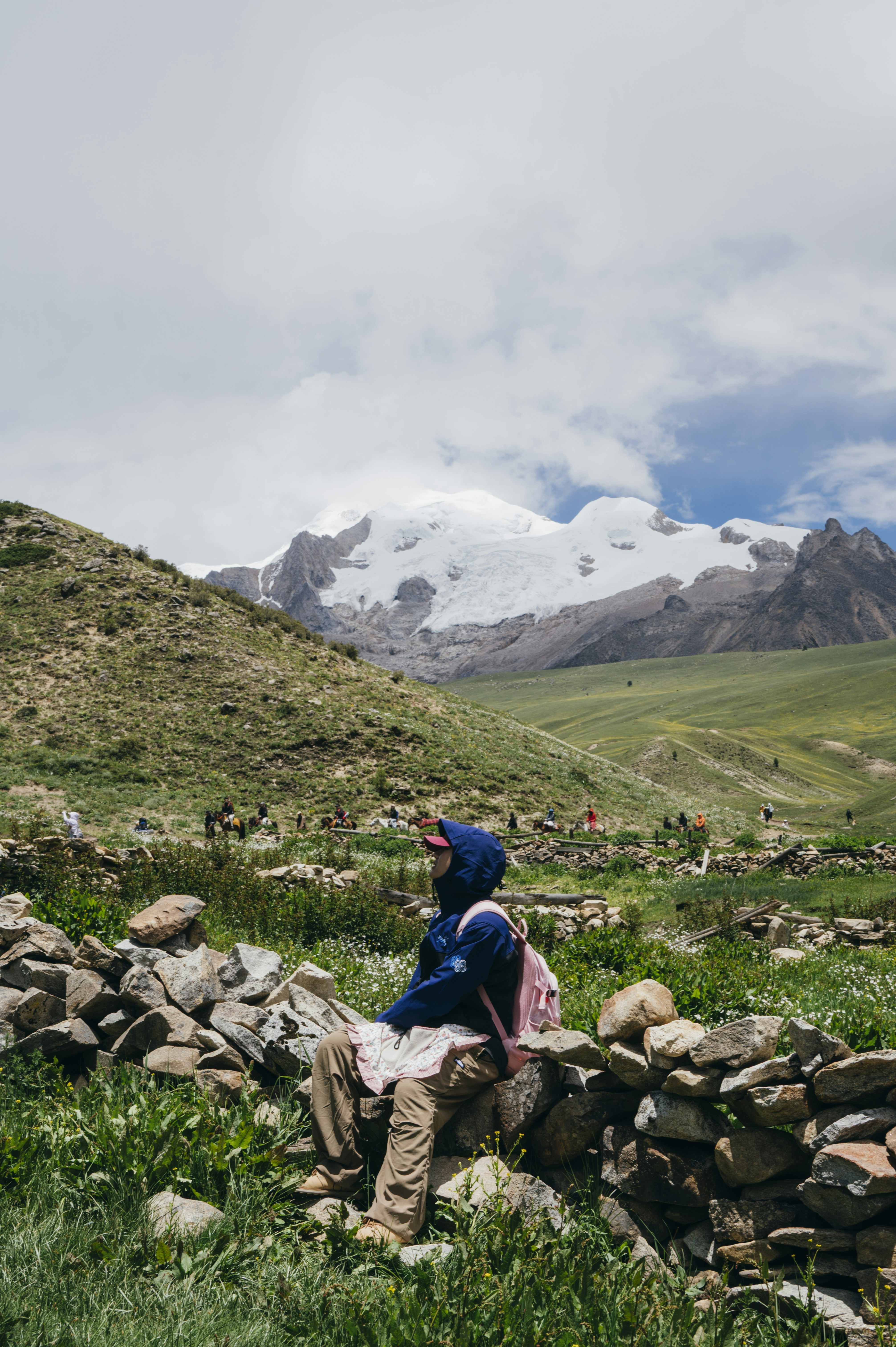 A hiker sits on a rustic stone wall in the foreground with a snowy alpine ridge in the distance.