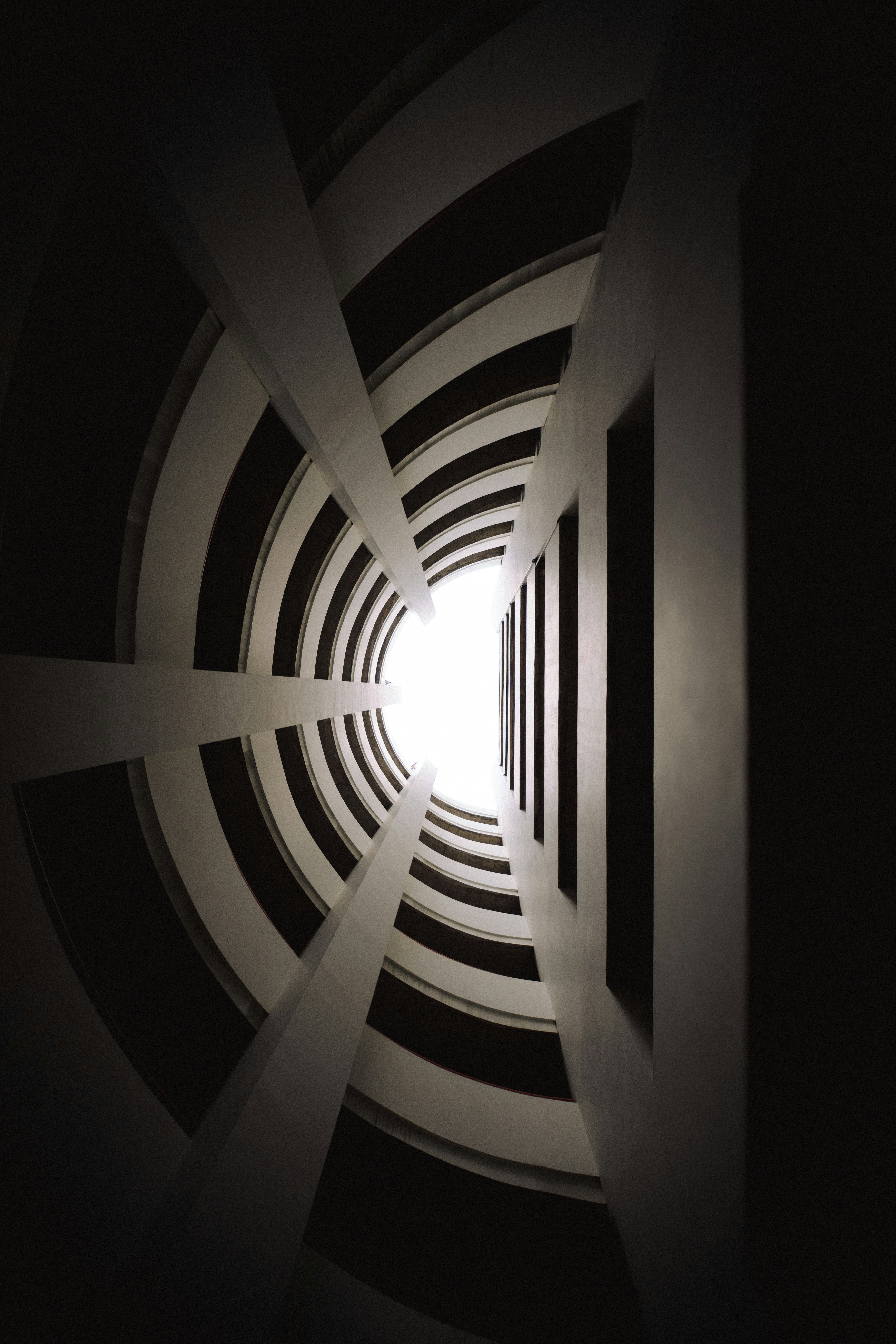A dark interior spiral leads the eye toward a bright central opening, created by concentric rings and angular supports. The image emphasizes geometry and depth, suggesting architectural confinement and a distant light source.