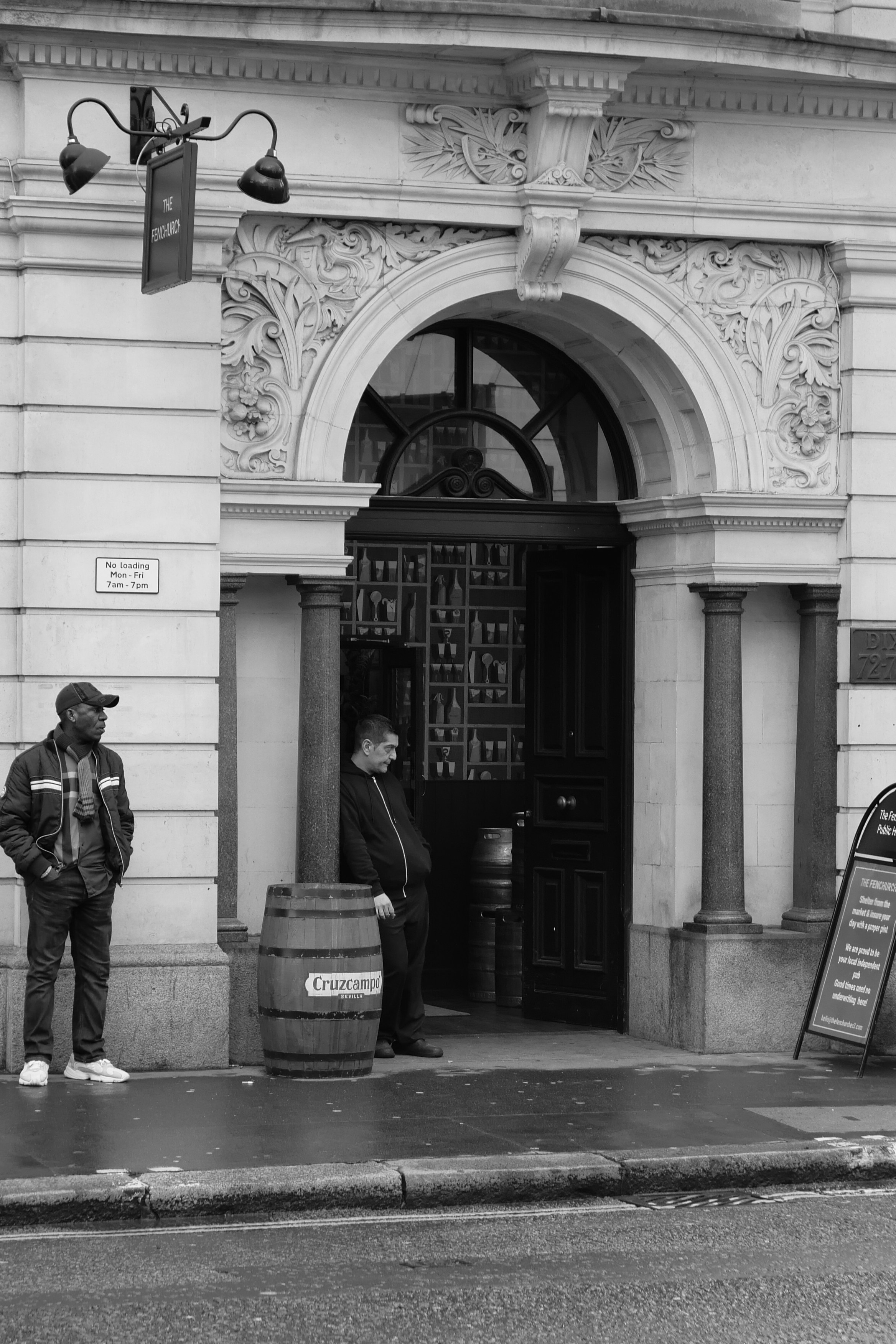 Monochrome street scene framed by an ornate building arch, with two men near a doorway and a wooden barrel in the foreground.