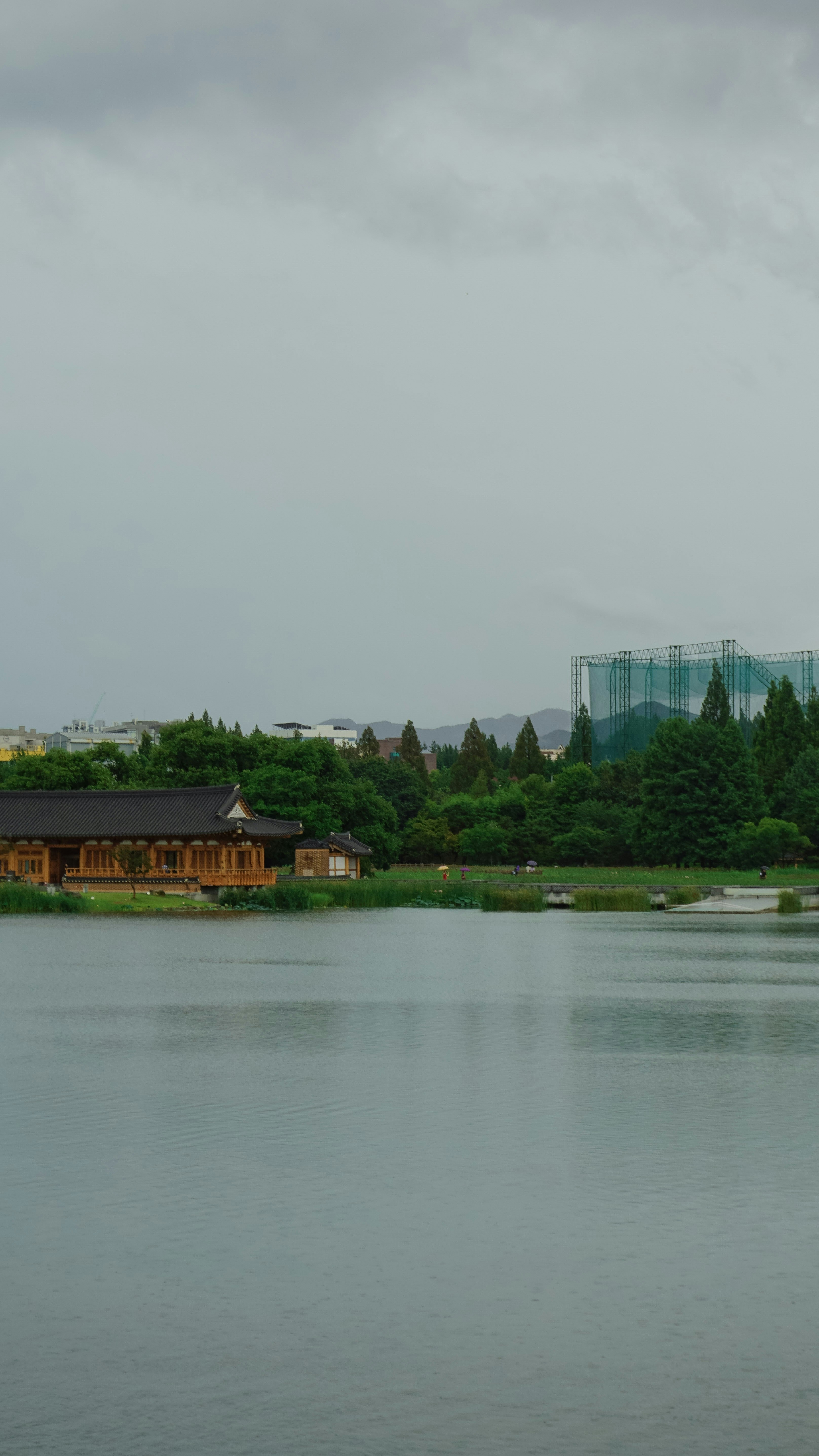 A tranquil lakeside scene featuring a traditional wooden pavilion along the shore, lush trees, and a distant structure under a pale, overcast sky.