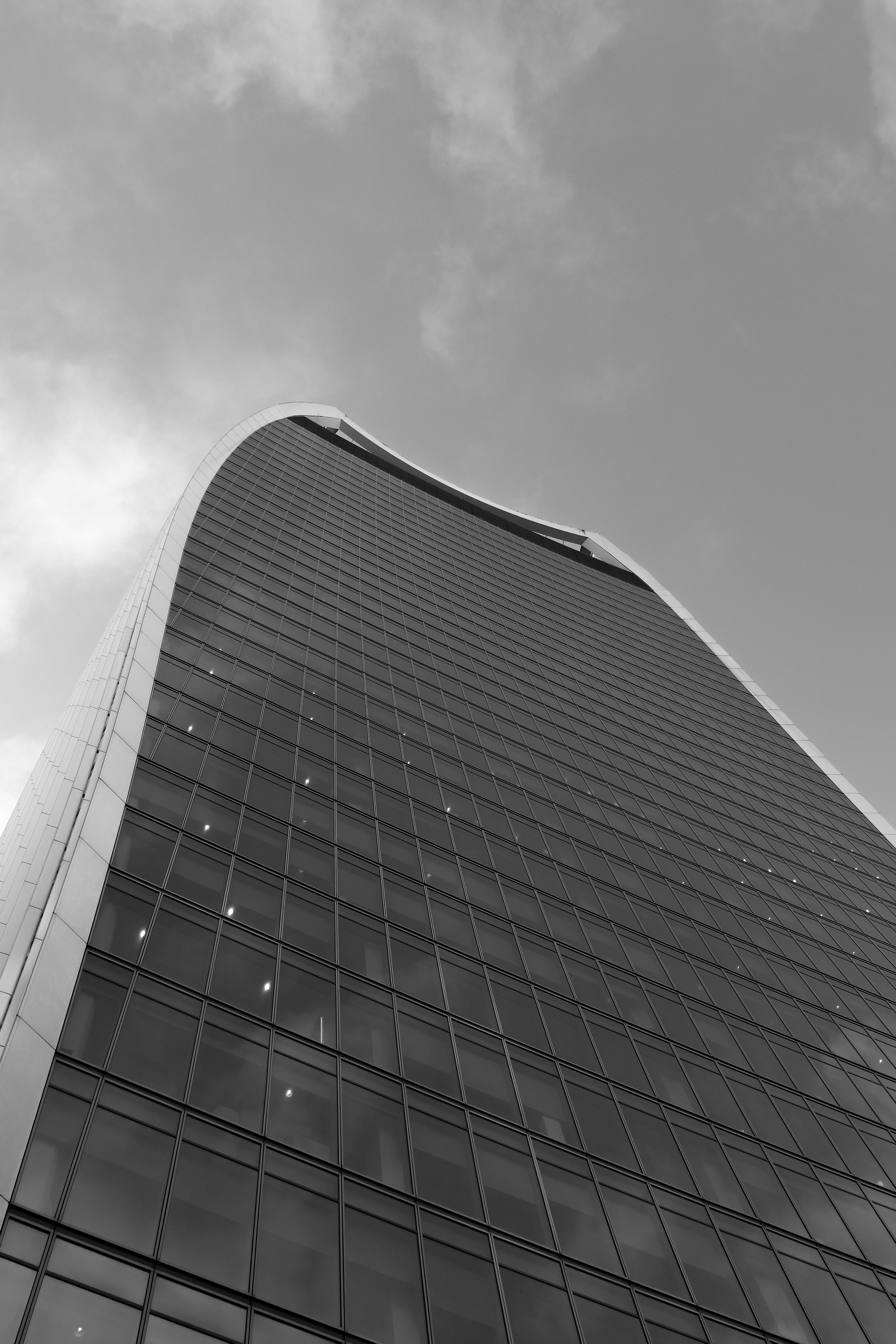 Sleek glass skyscraper photographed from a low angle, emphasizing its curved silhouette against a cloudy sky. A modern architectural study focusing on reflection and structure.