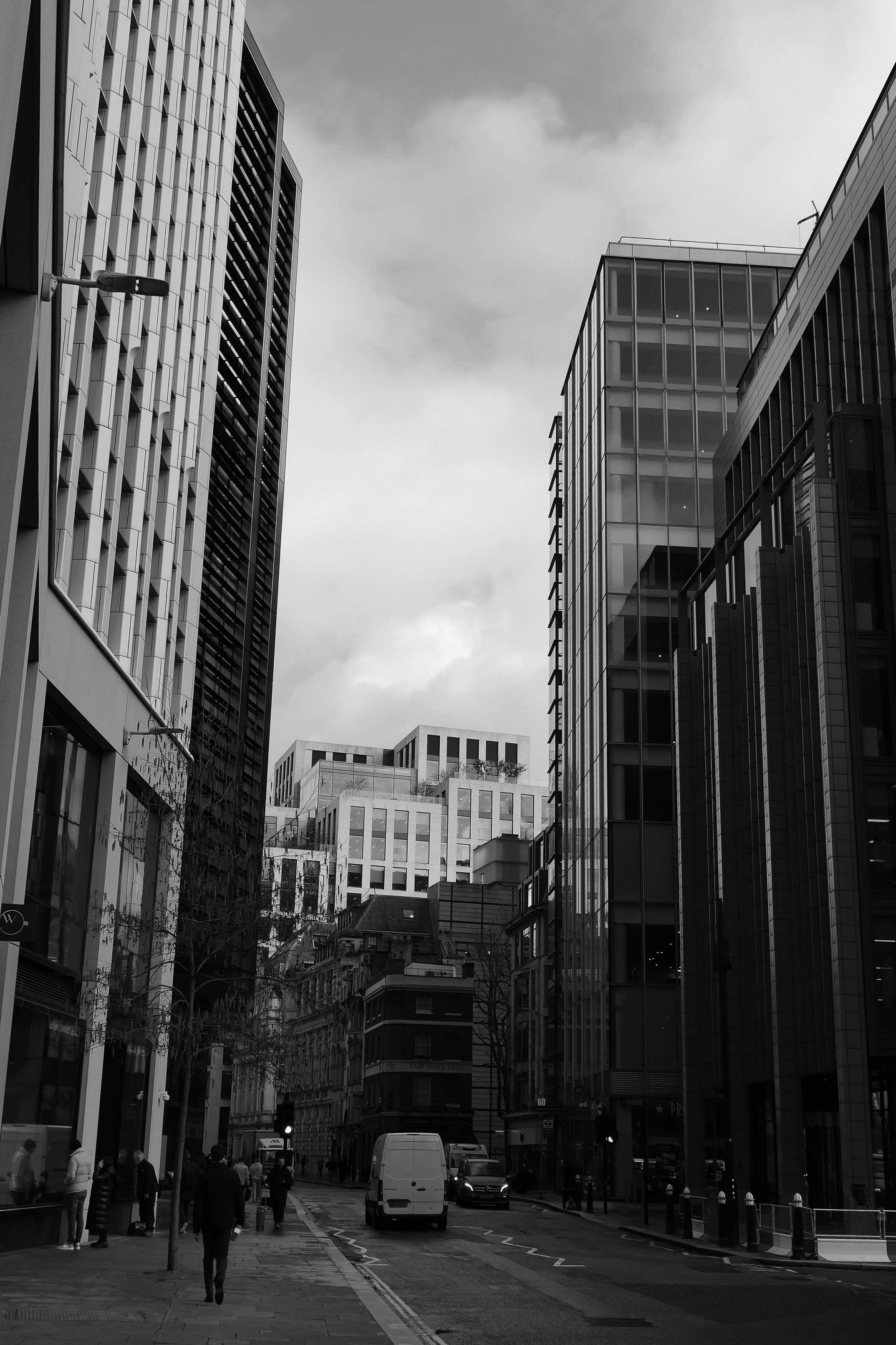 Monochrome urban street scene framed by tall glass and concrete buildings, with pedestrians and a van along a narrow avenue.