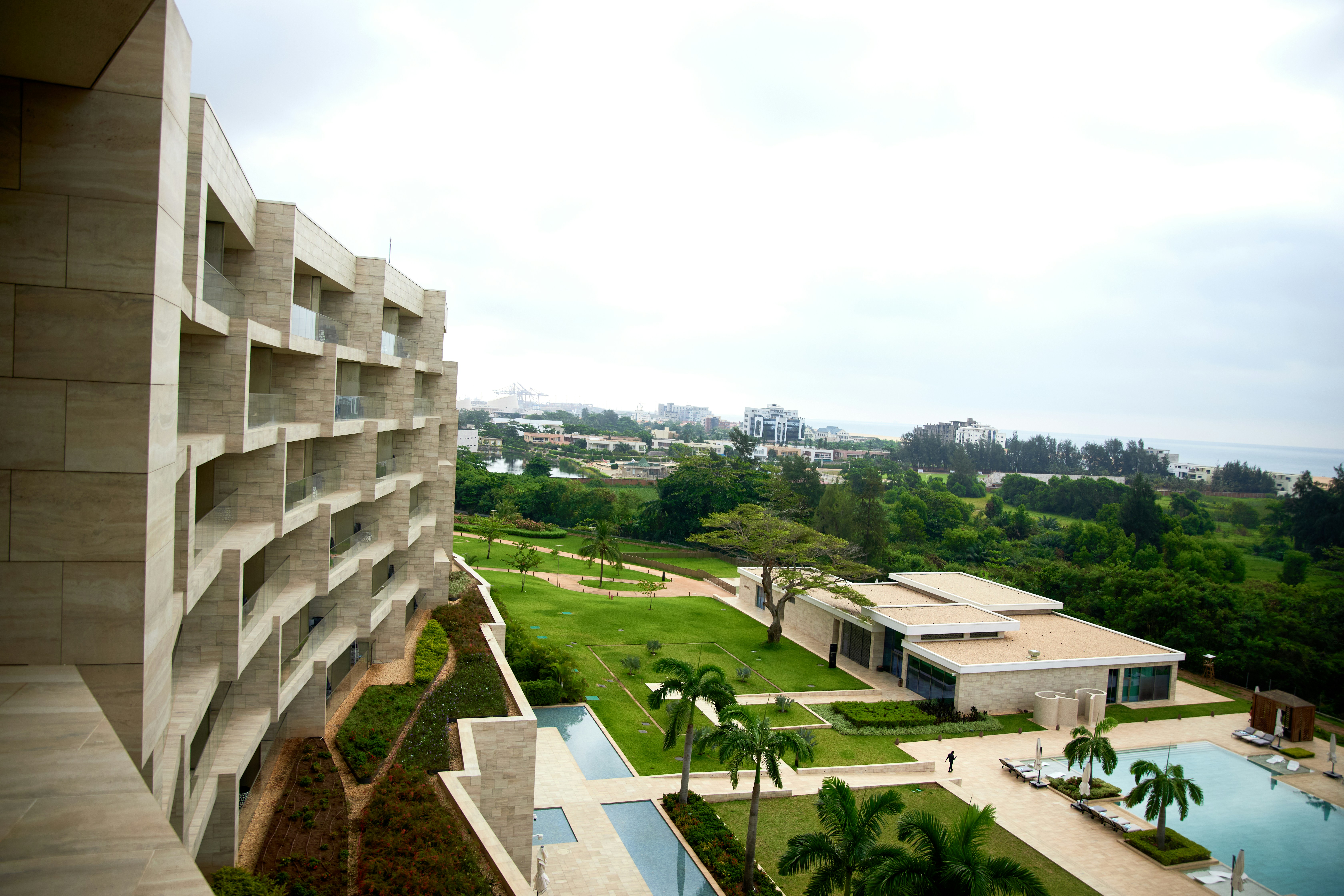 Wide-angle view of a modern architectural complex with a textured facade on the left, green lawns, palm trees, and a pool area extending toward a distant cityscape.