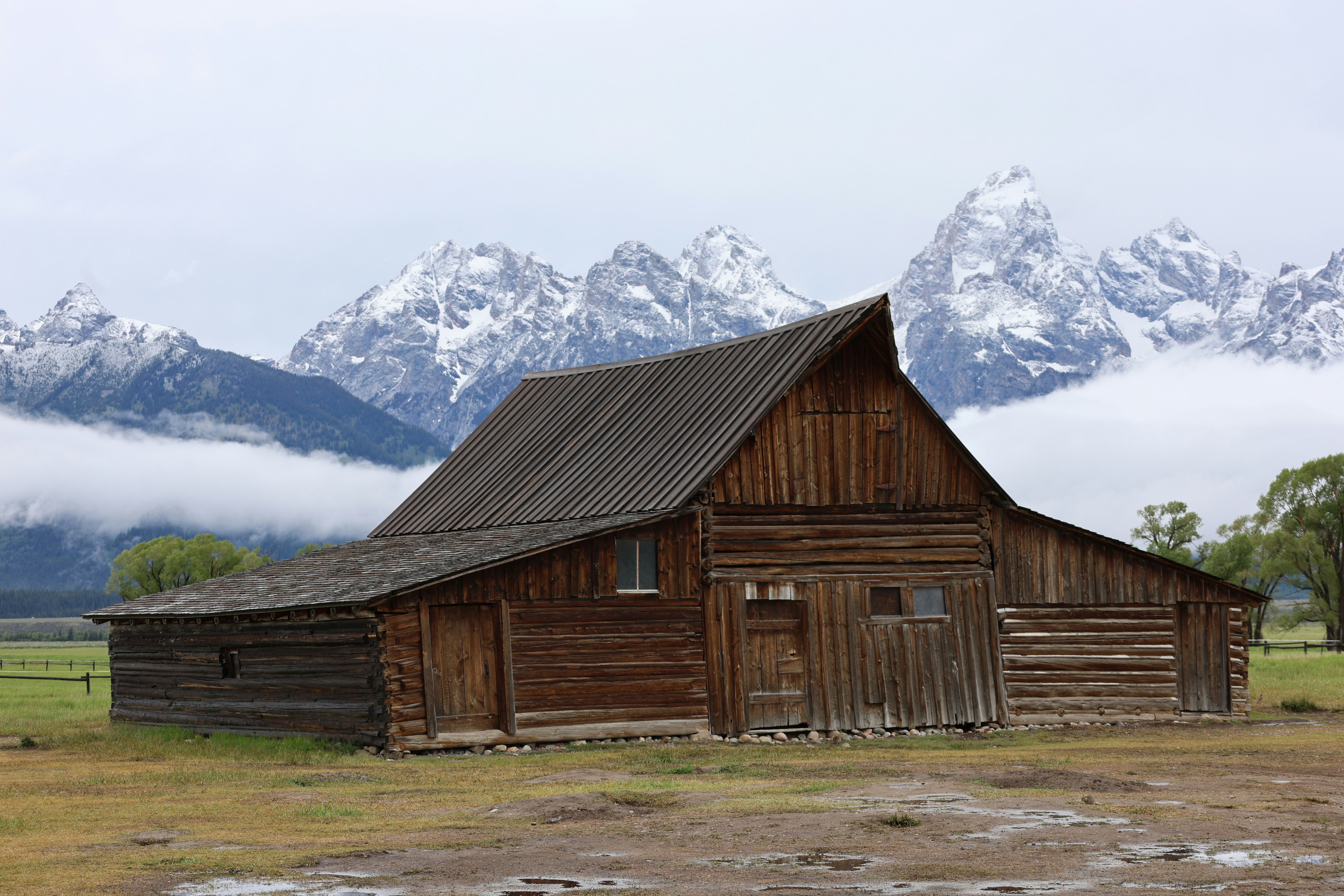 A weathered wooden barn sits in a grassy field with a dramatic alpine backdrop of snow-capped mountains and low-lying clouds.