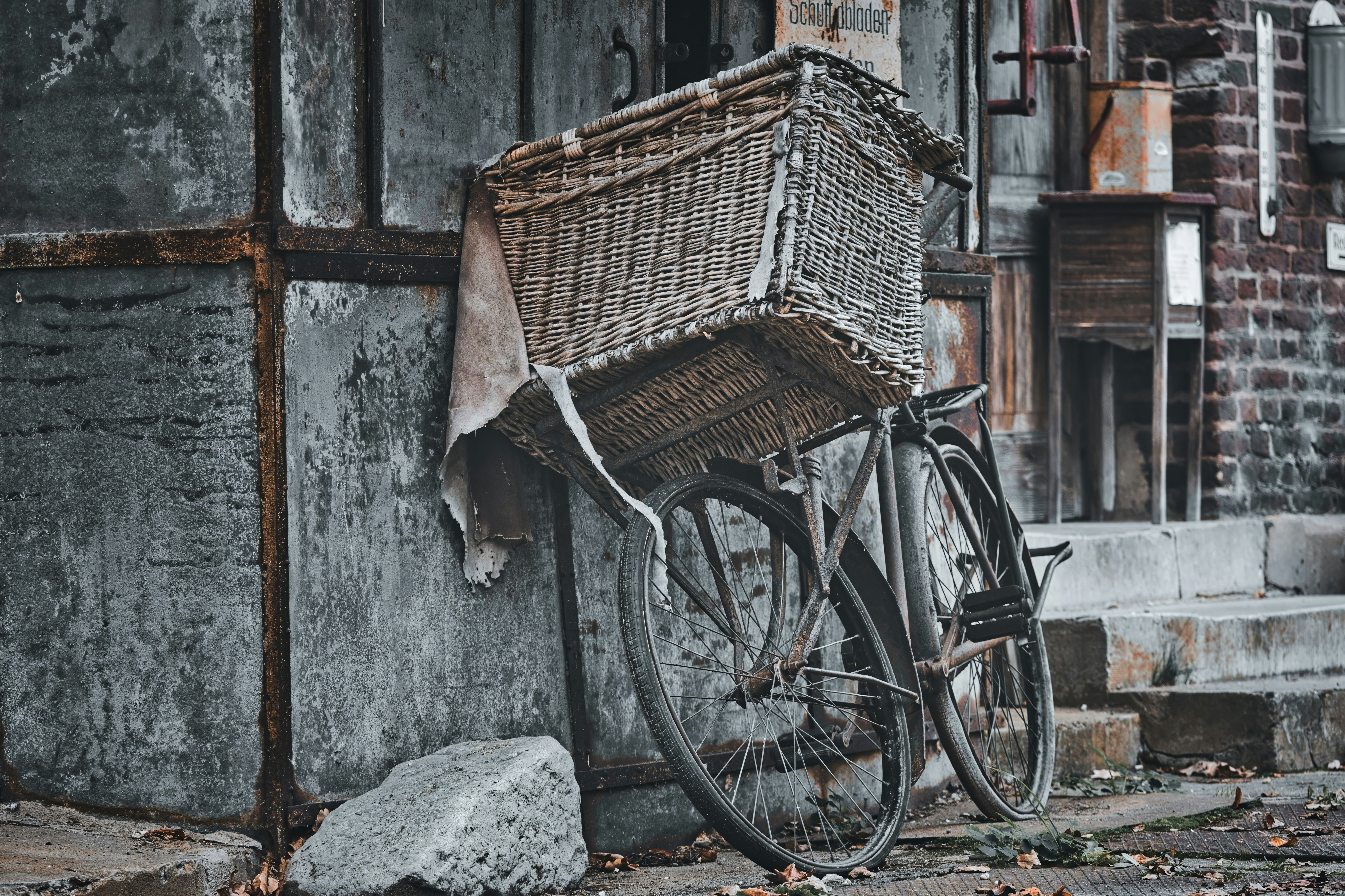 A weathered bicycle with a woven basket rests against a brick wall, dusting the ground with fallen leaves and rough textures of urban neglect.