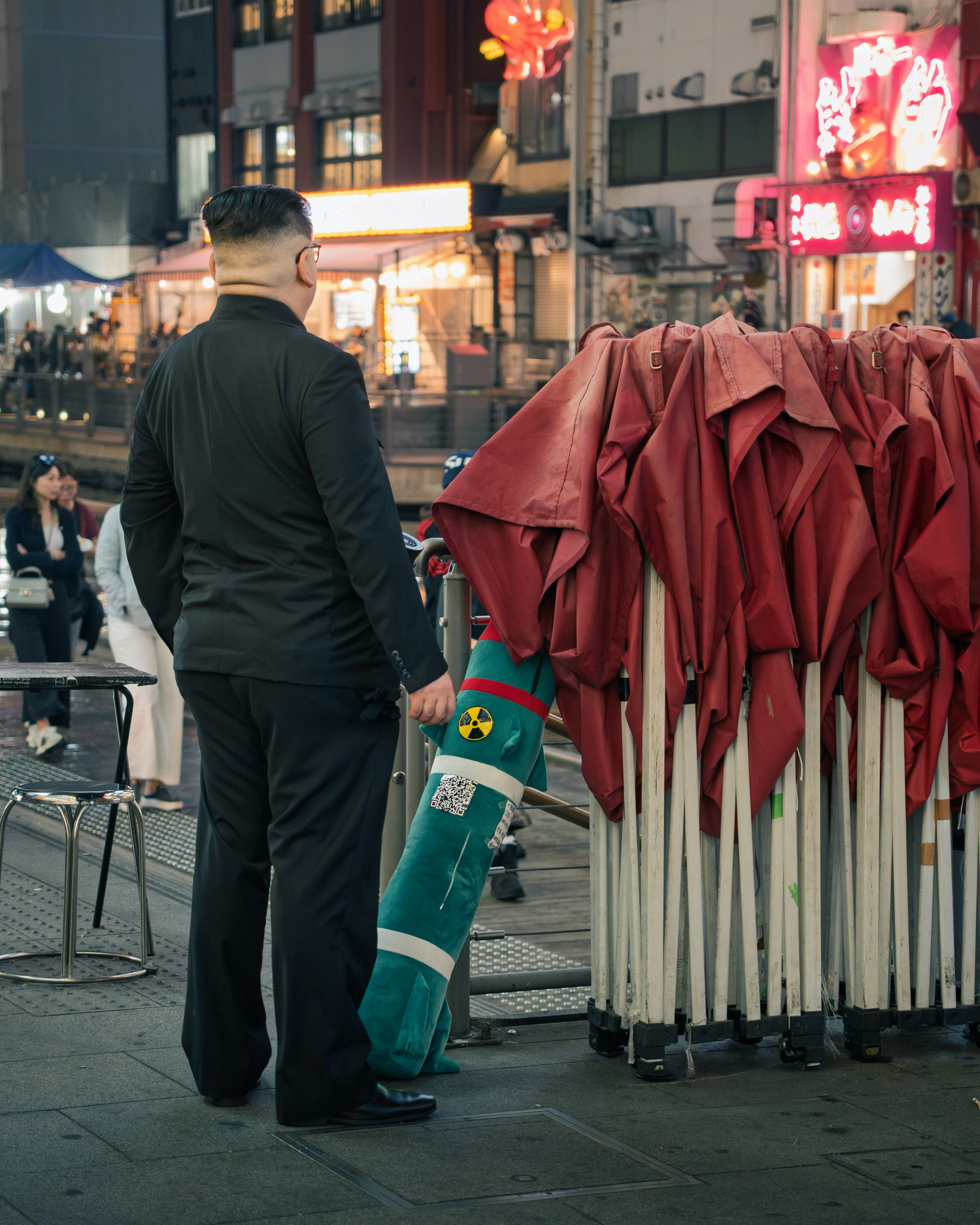 Urban street scene with a man in a black suit standing near red umbrellas on a city sidewalk at dusk.