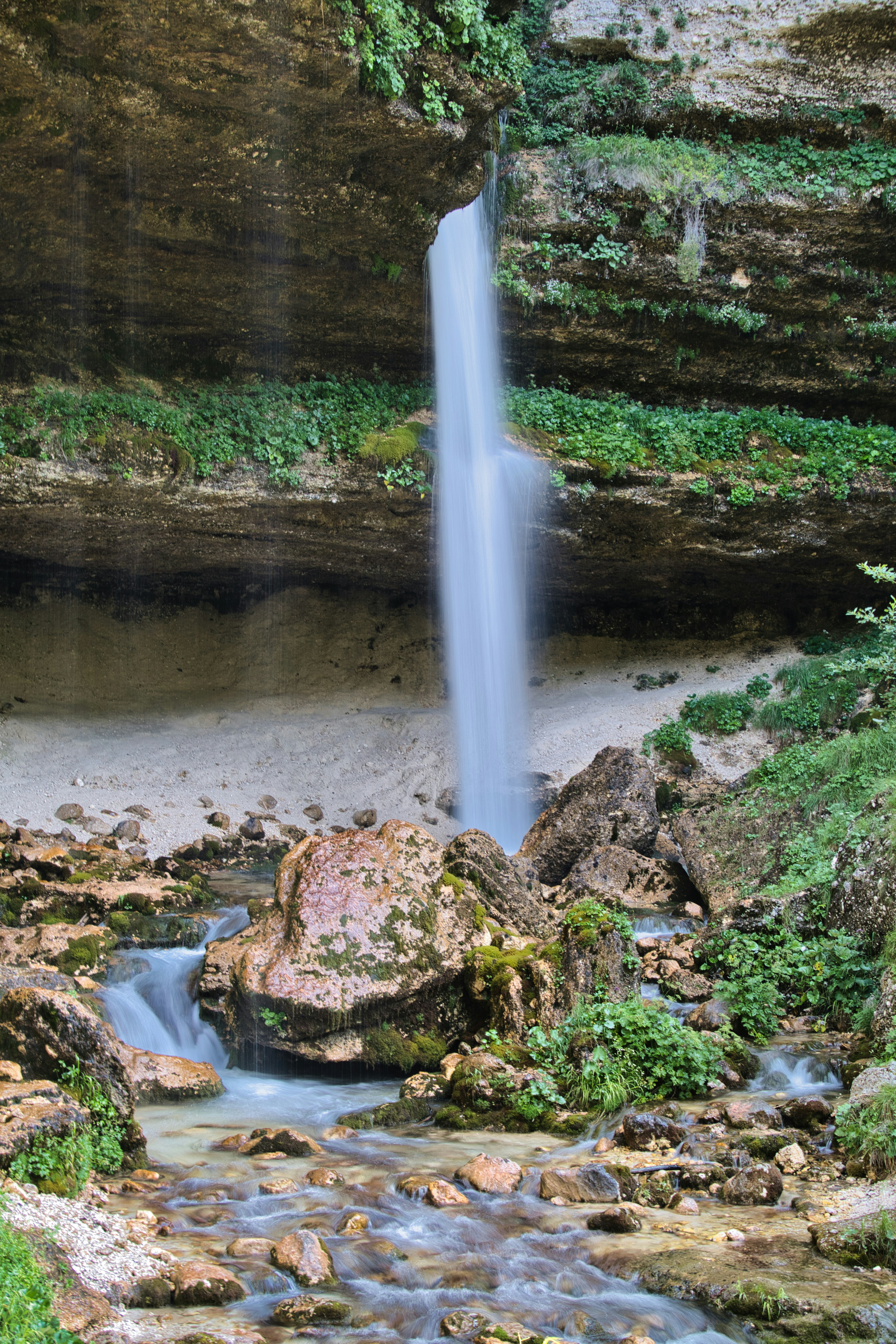 Tall waterfall spilling from a rock overhang into a rocky pool below, framed by green foliage and layered cliff walls.