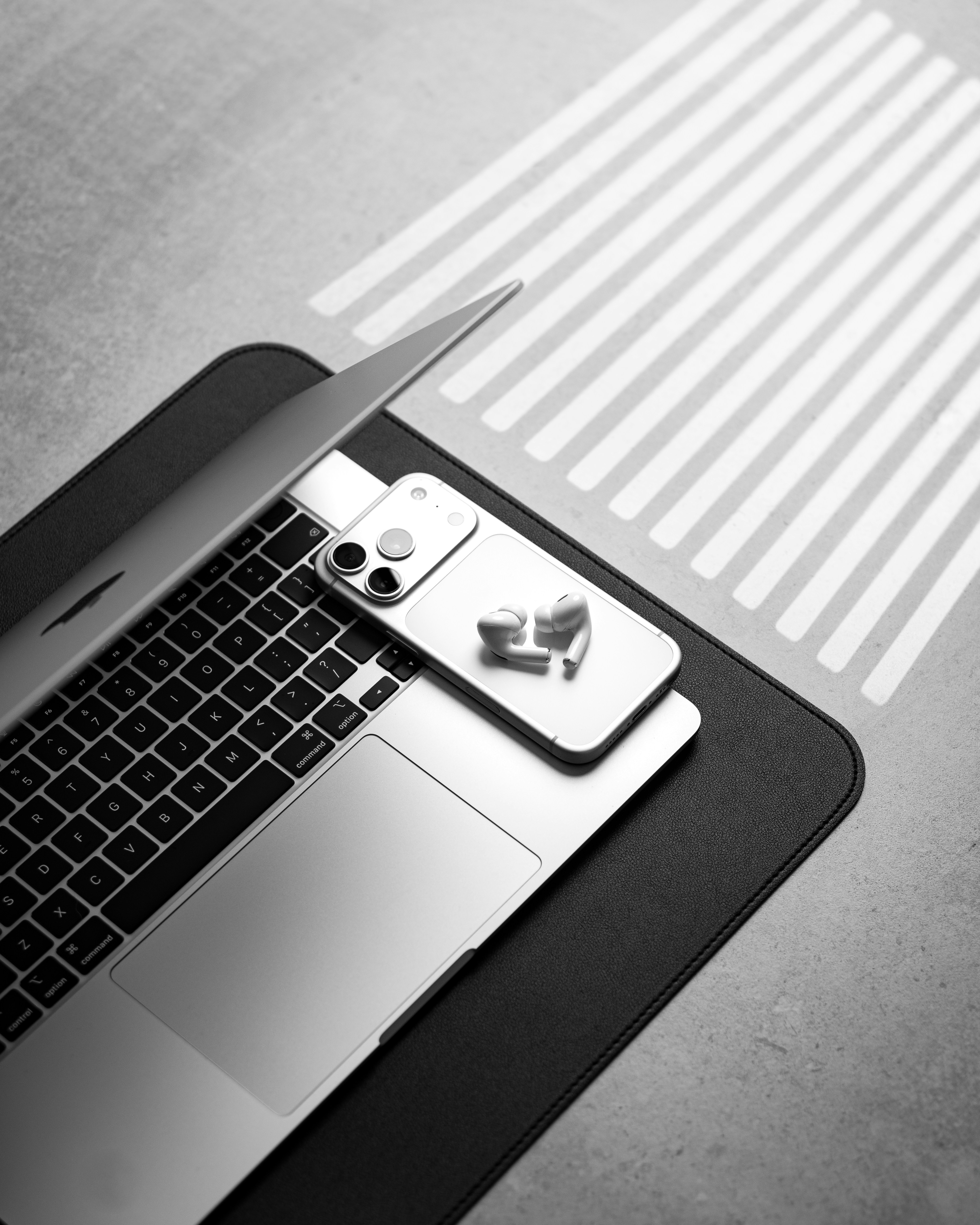 A silver laptop with wireless earbuds sits on a black sleeve atop a desk, angled under an open lid with a concrete surface and striped crosswalk motif in grayscale.
