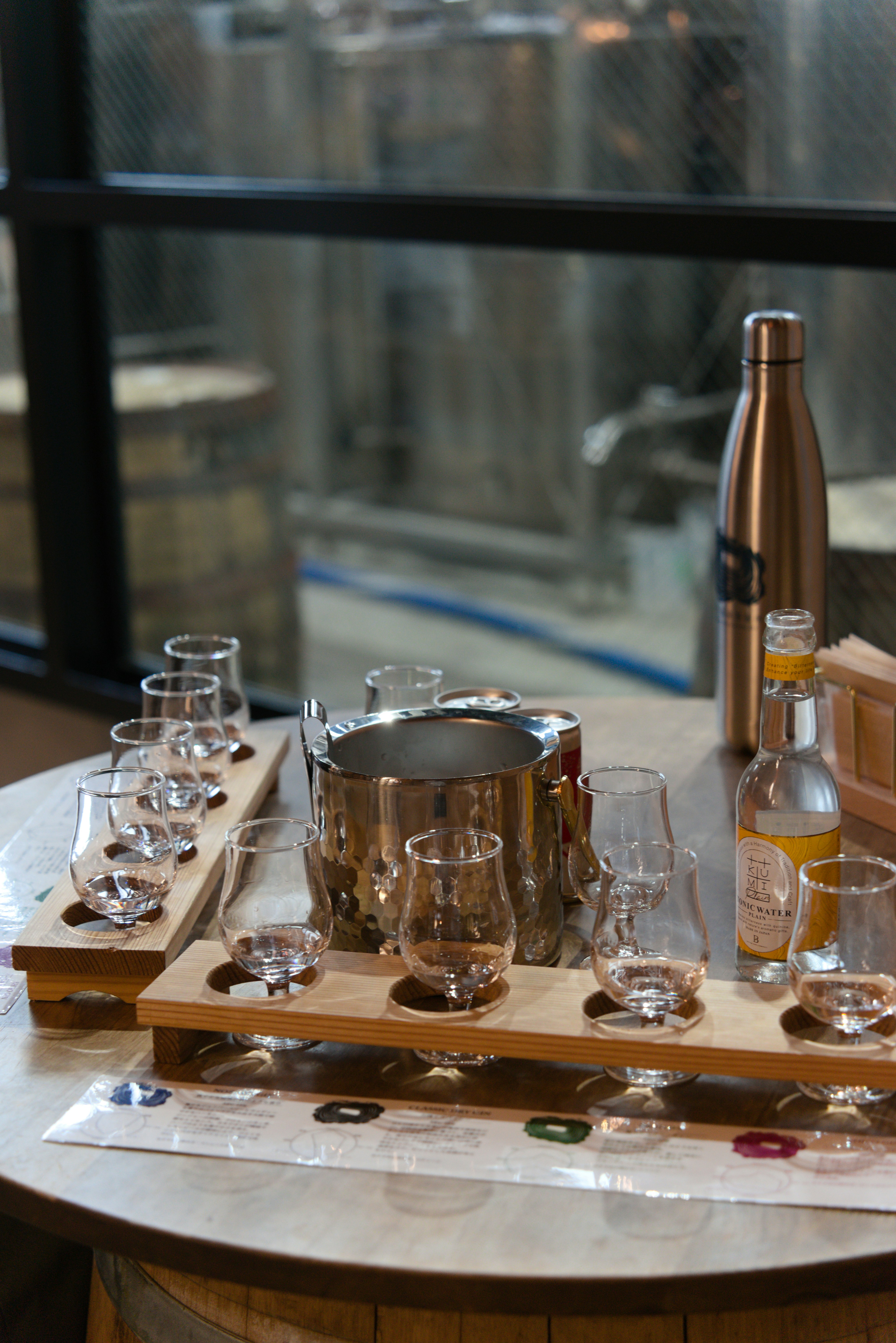 Close-up view of a wooden beverage tray with multiple glassware pieces and a metal ice bucket on a rounded bar table; a bottle and a bottle of spirits sit in the background by a large window.