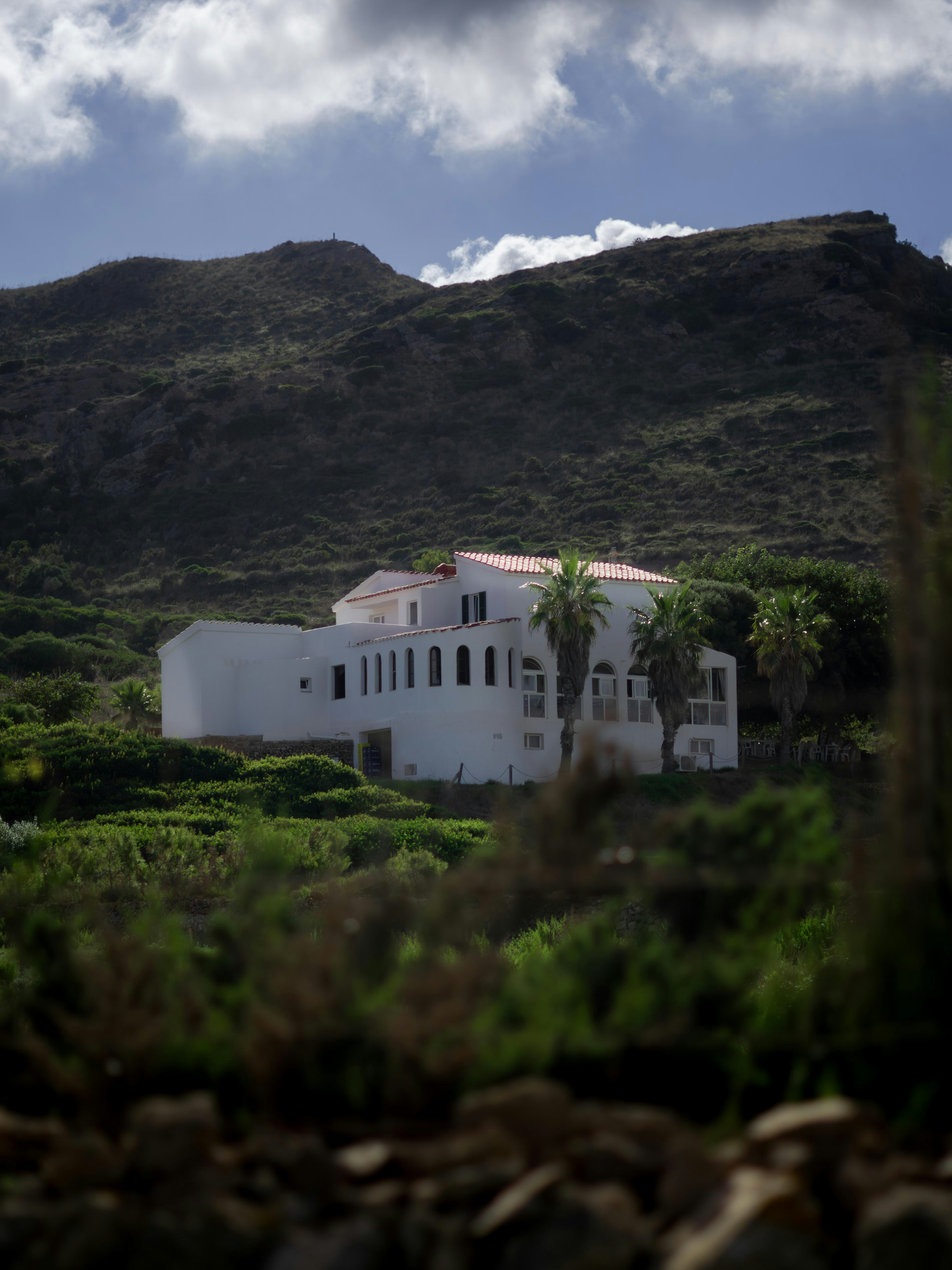 A bright white villa with arches sits in lush greenery at the base of rugged hills under a blue sky. The scene emphasizes Mediterranean architecture against natural terrain.