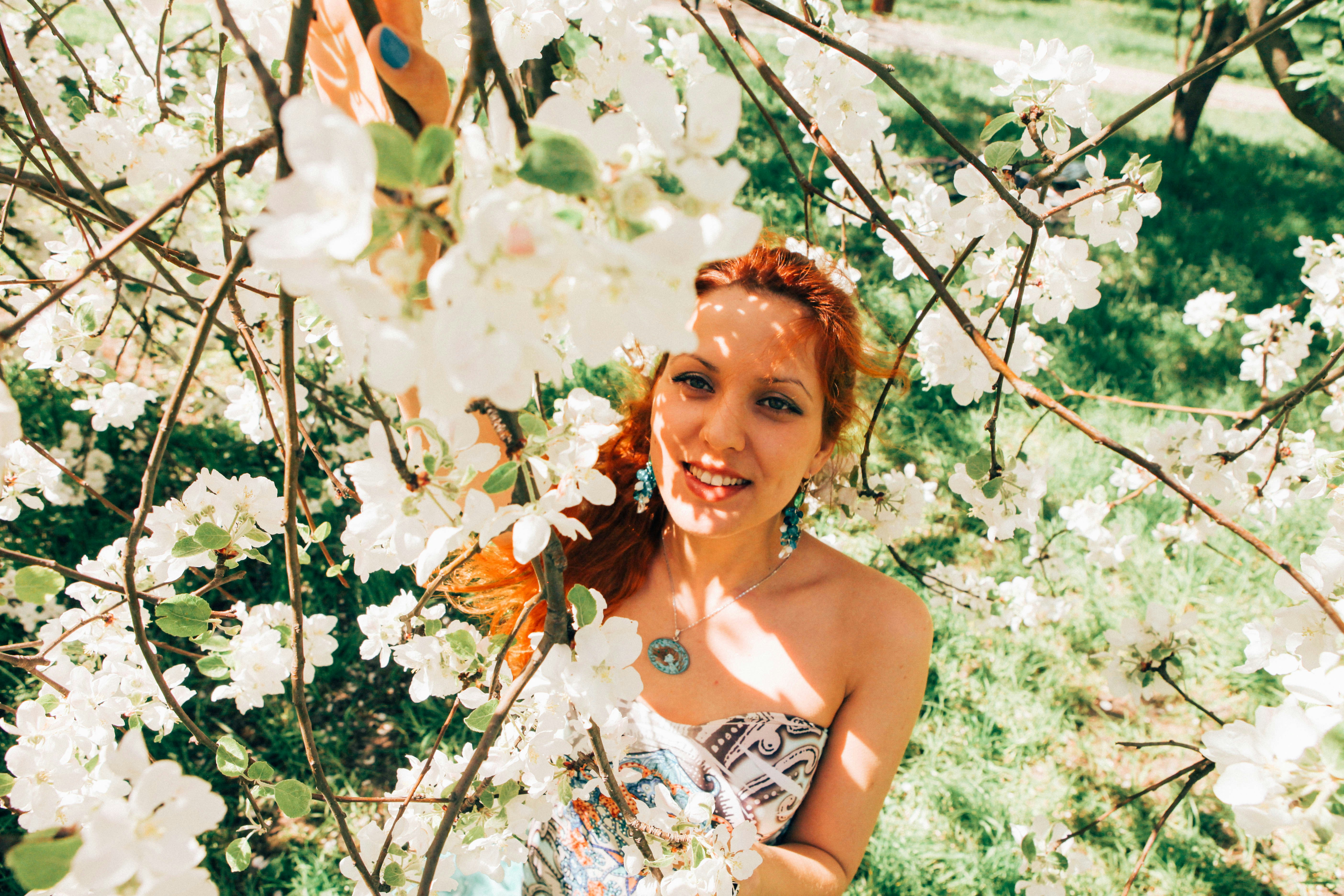 A smiling woman stands among white blossoms in bright garden light, framed by flowering branches. The scene highlights a warm, candid portrait with natural floral backdrop.
