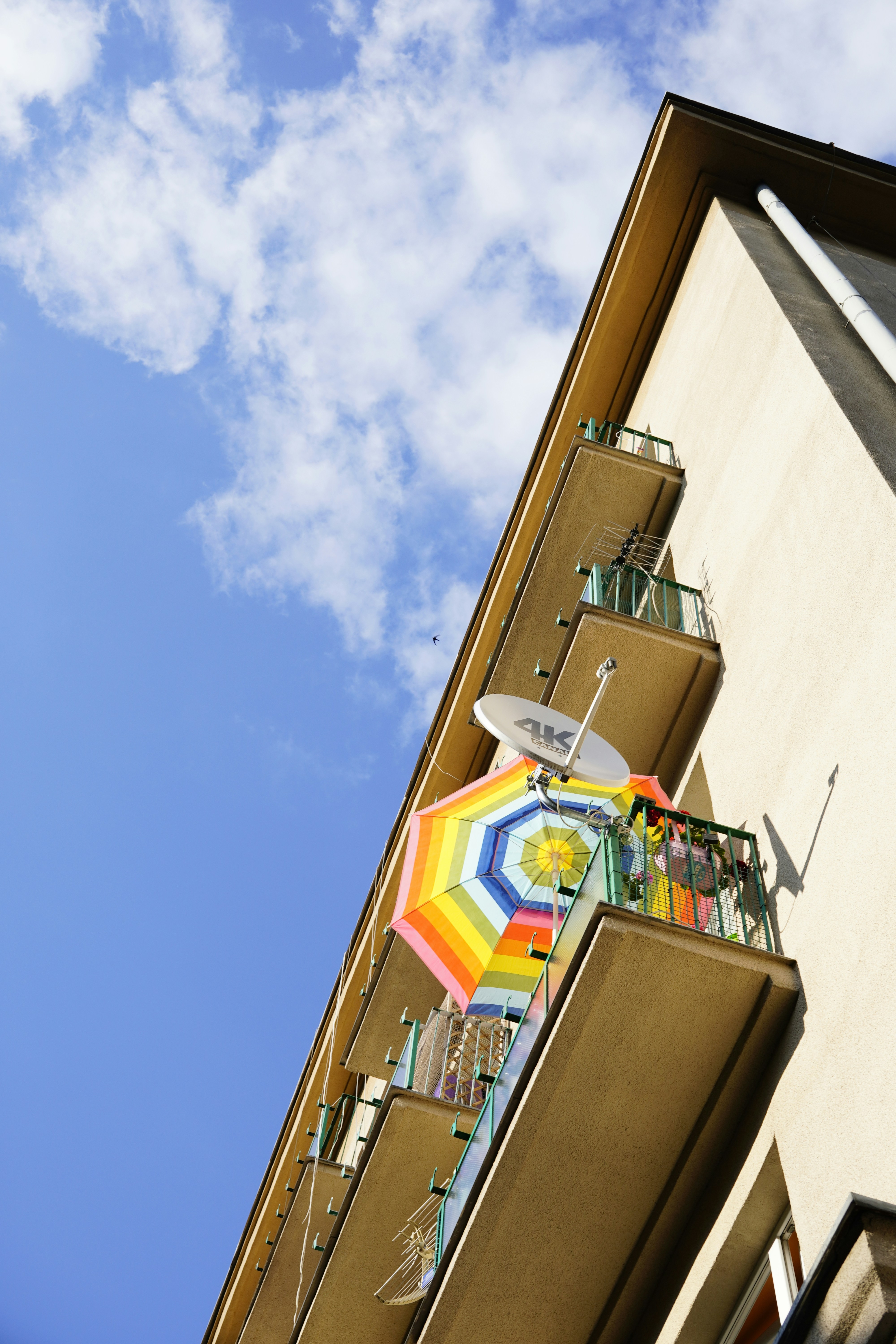 Bright rainbow umbrella on a mid-rise balcony against a clear blue sky and beige building facade.