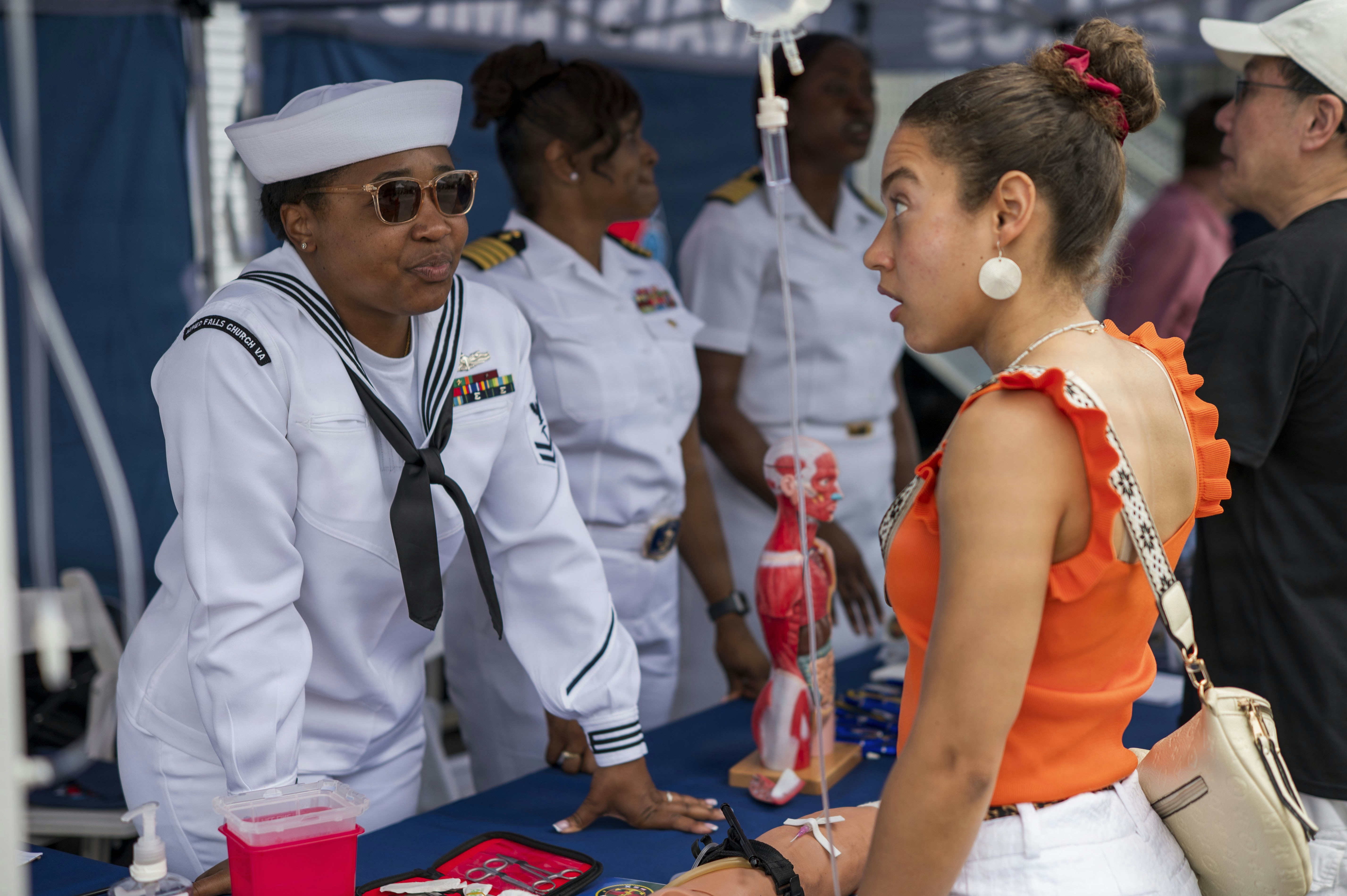 A Navy officer in uniform converses with a young dancer at a booth, with other naval personnel and onlookers in the background; the scene appears to be at a public outreach event or festival.