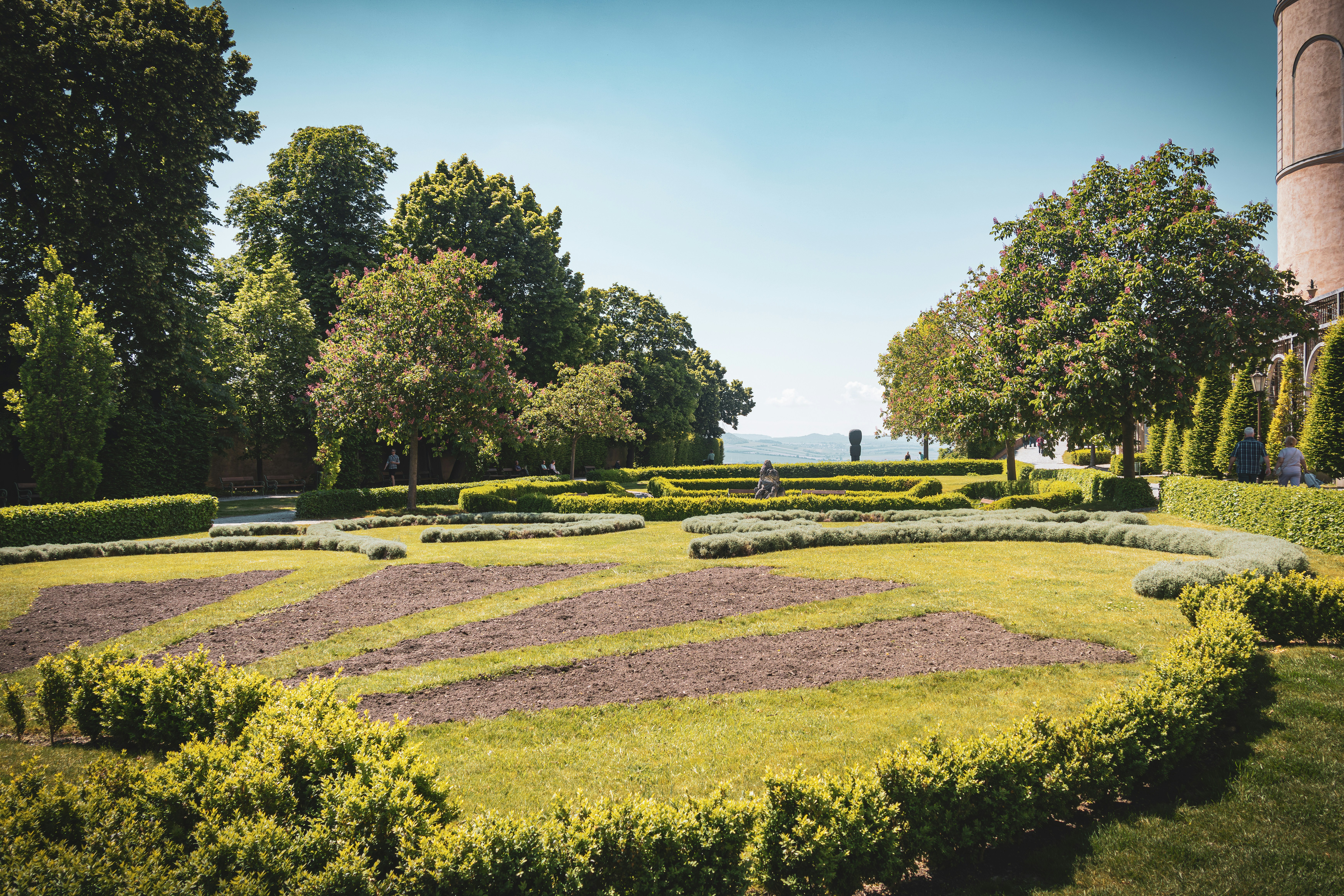 Vibrant formal garden with layered hedges and circular patterns leading to a distant horizon; a tall architectural element rises on the right, under a clear sky.