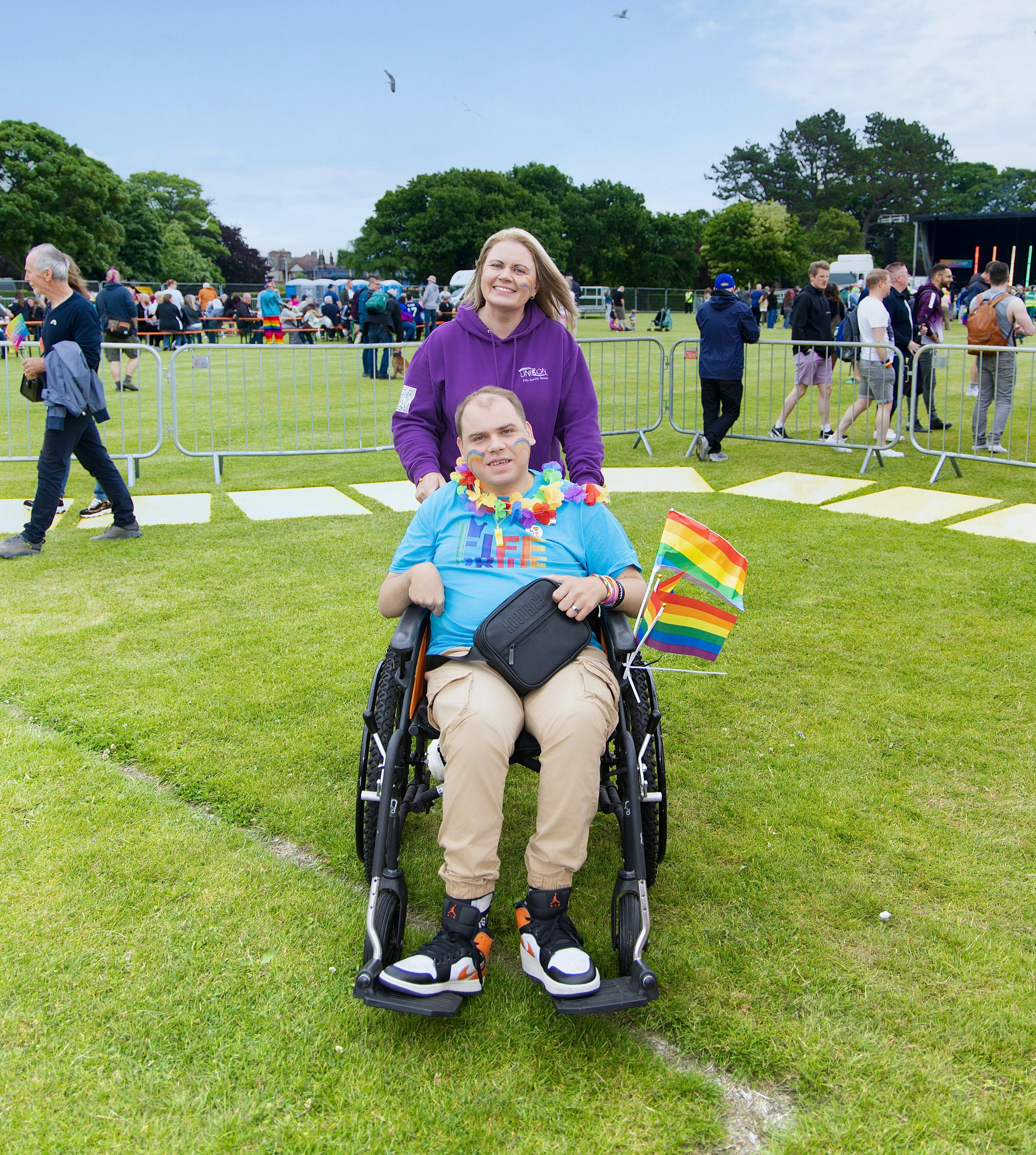 Man in wheelchair rides toward camera with rainbow flag, accompanied by a smiling helper; crowd and event barriers in the background at a sunny park.