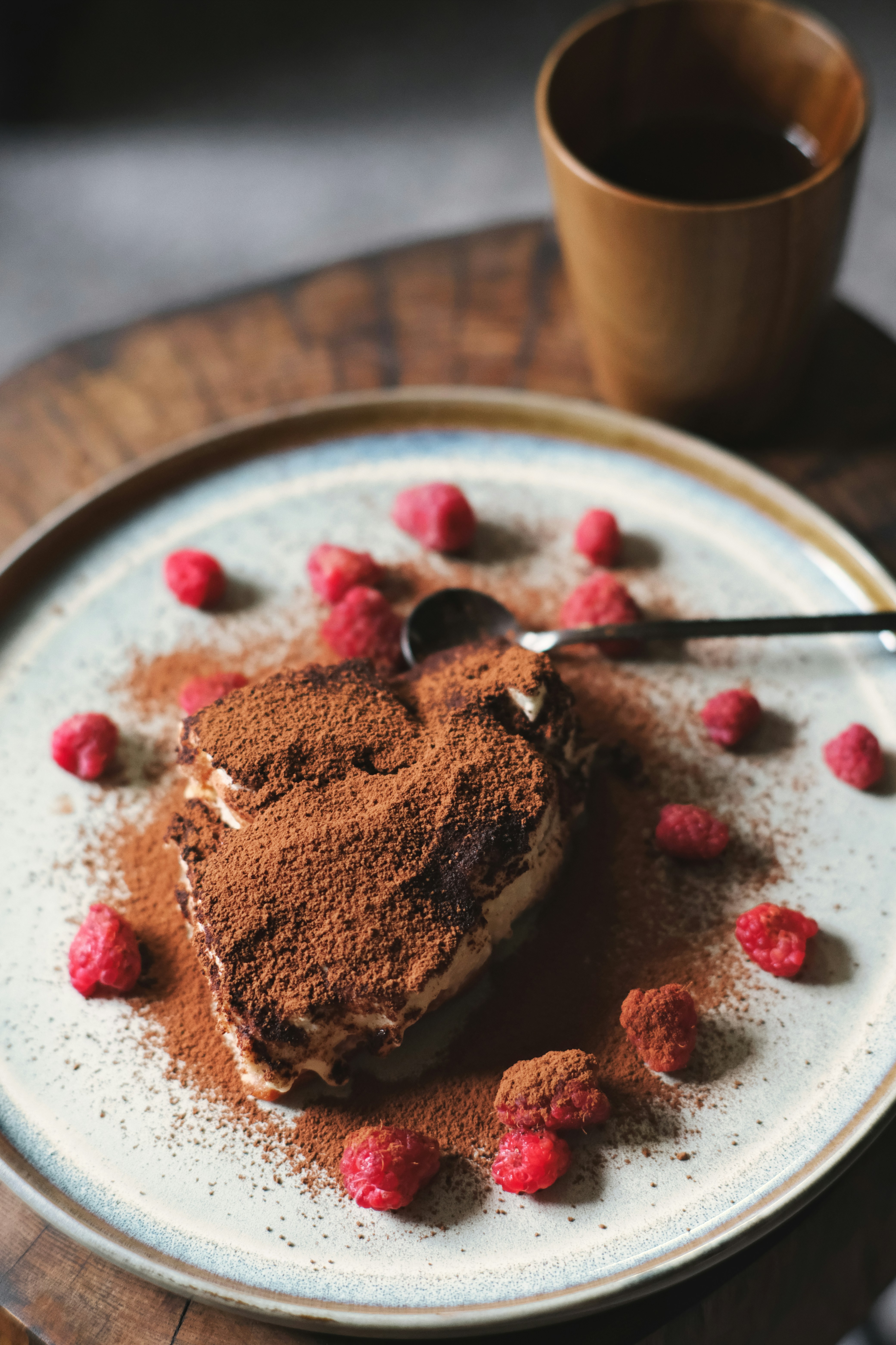 Chocolate dessert on a frosted plate with cocoa powder and raspberries, accompanied by a wooden cup in the background.