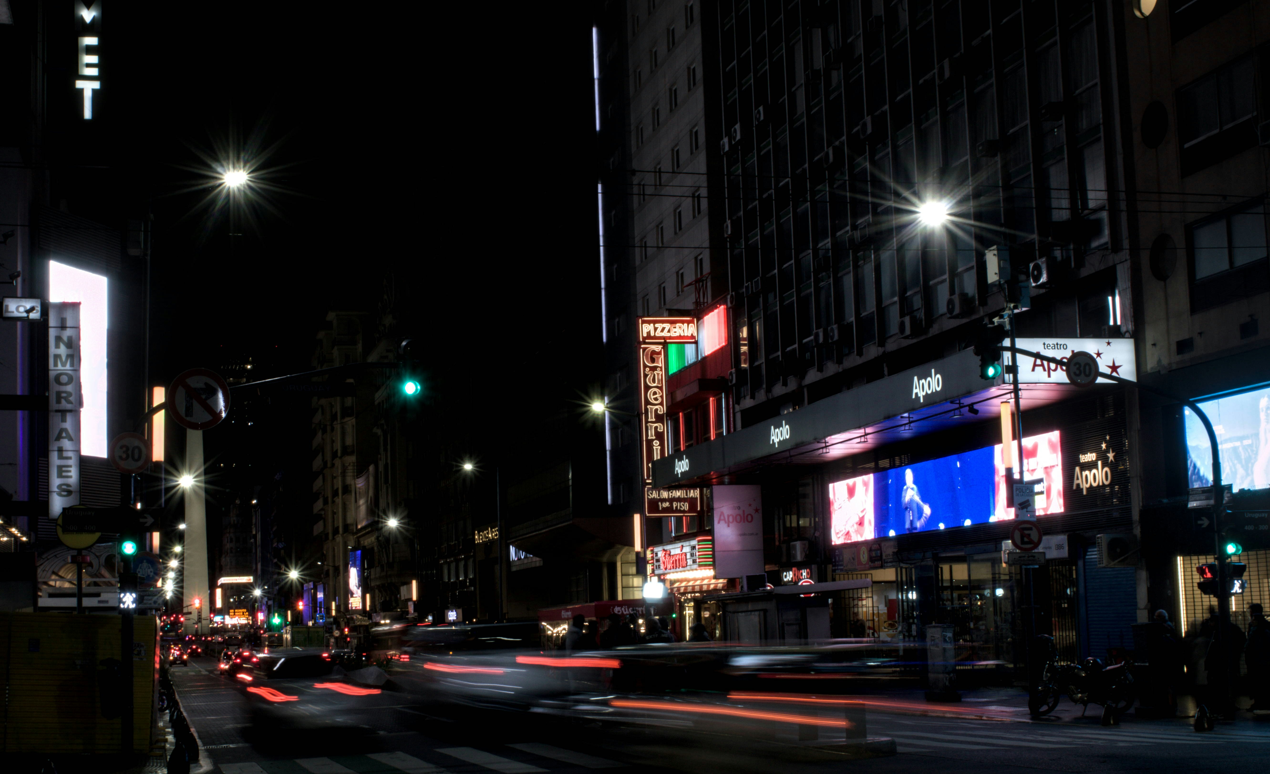 Bustling city street at night with motion trails of vehicles and illuminated storefronts; a long exposure captures light streaks along the avenue.
