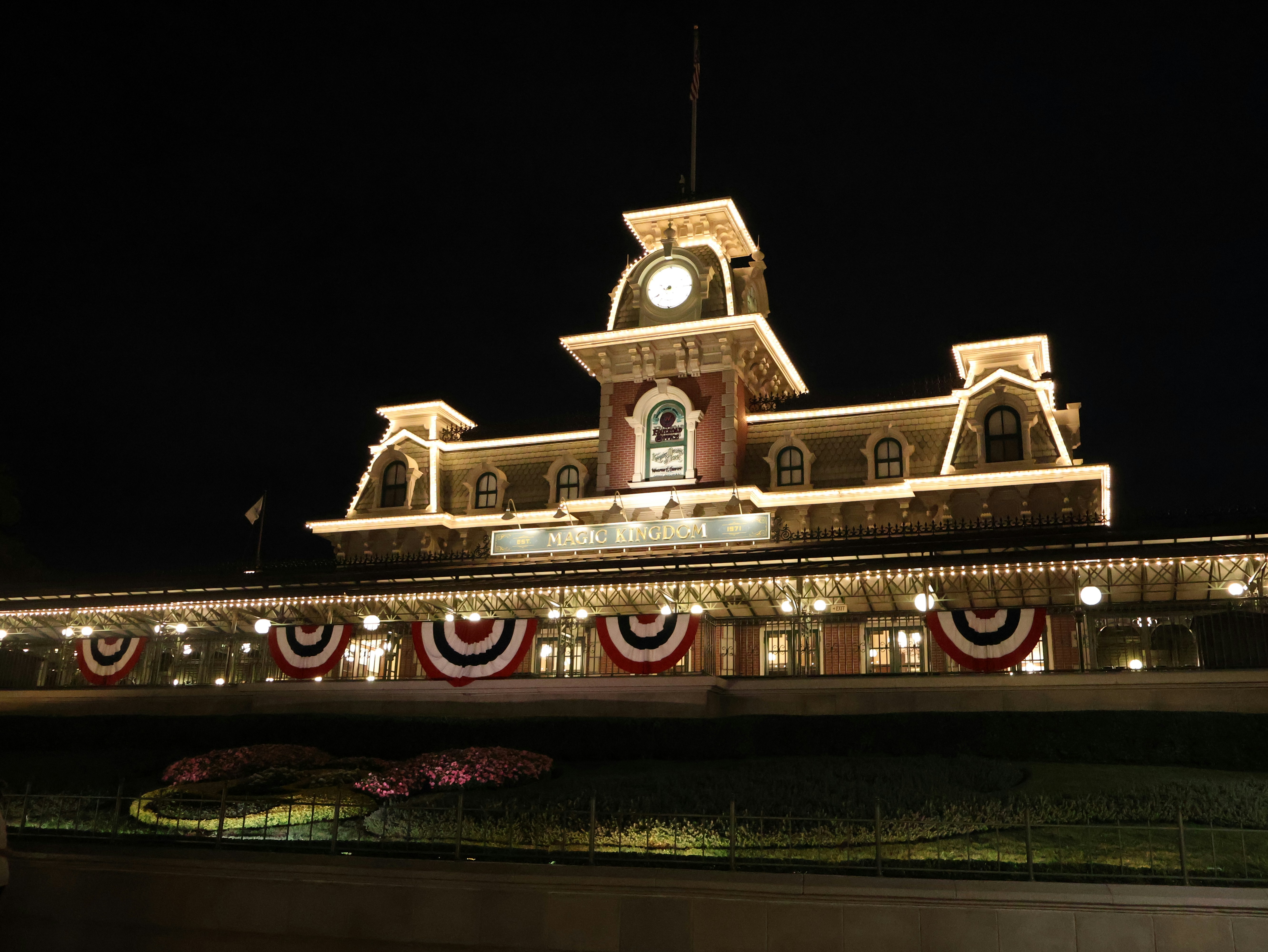 Night scene of a lit train station facade with a central clock tower, garlands, and bunting. A sense of holiday celebration, showing architectural detail and warm illumination.