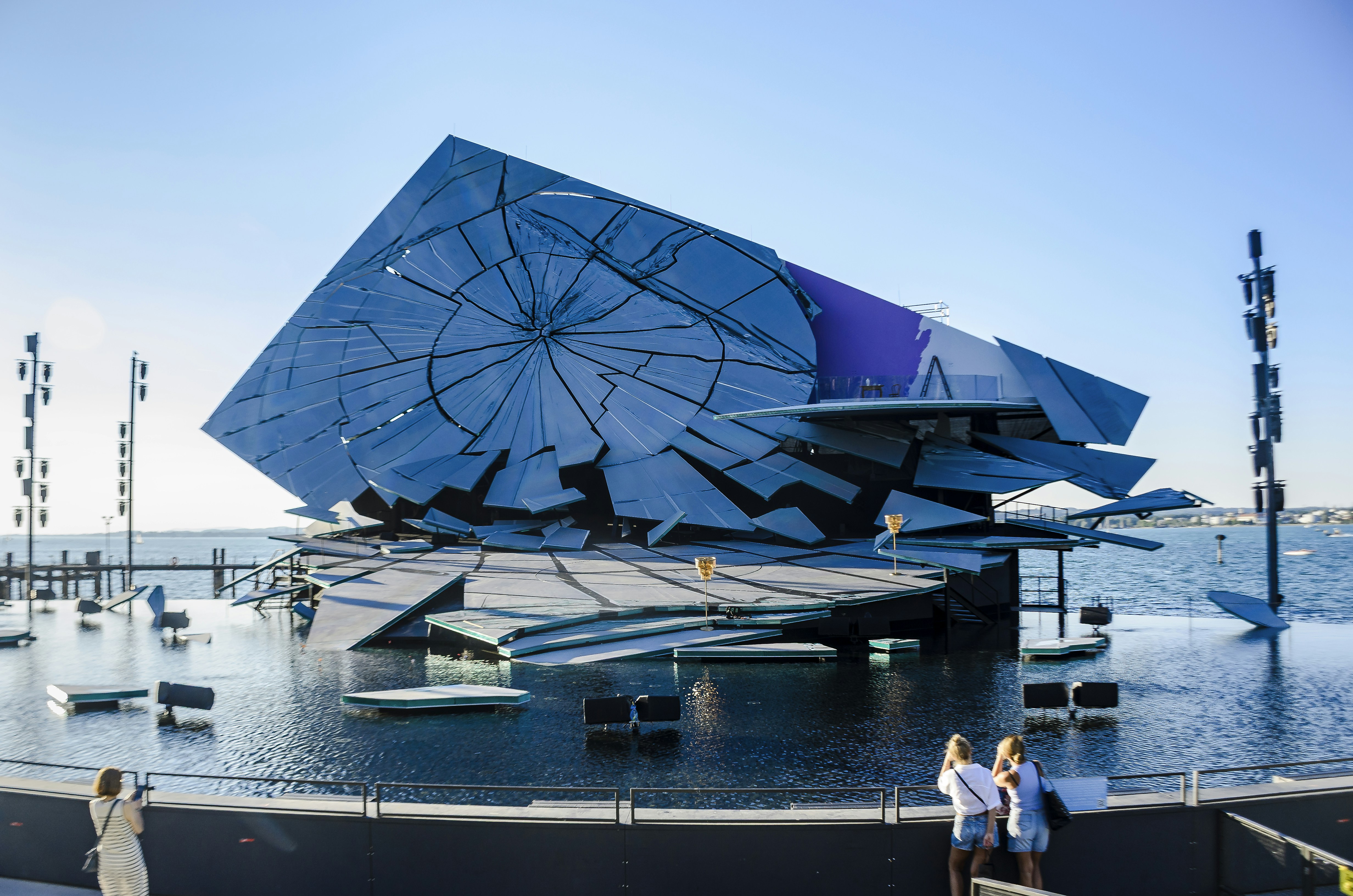 Blue angular sculpture dominates a calm harbor, with small boats and a railing in the foreground and a clear blue sky above.