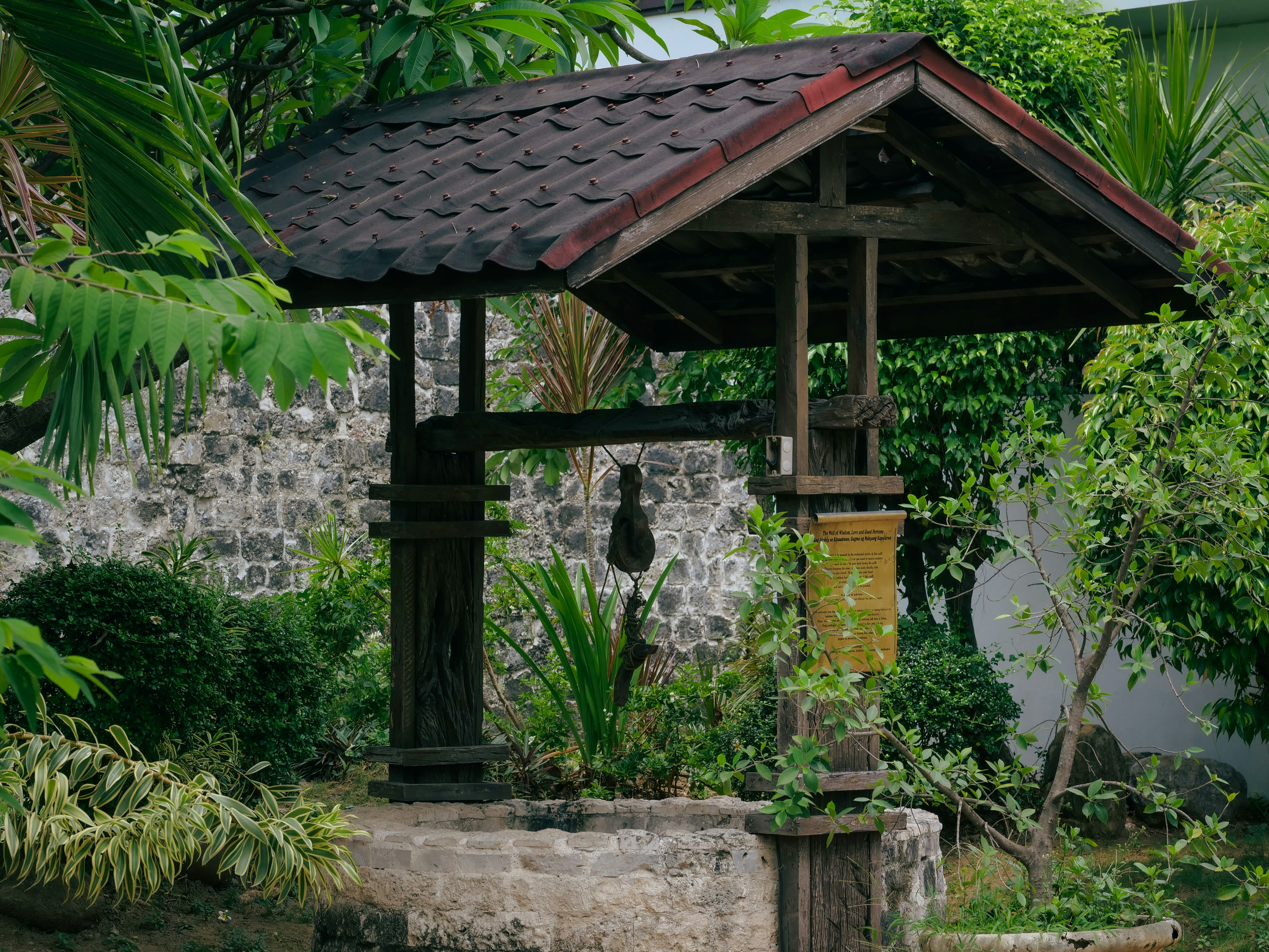 A small wooden shrine on a stone plinth sits within a tropical garden bordered by a mossy wall and dense foliage, creating a tranquil courtyard scene.