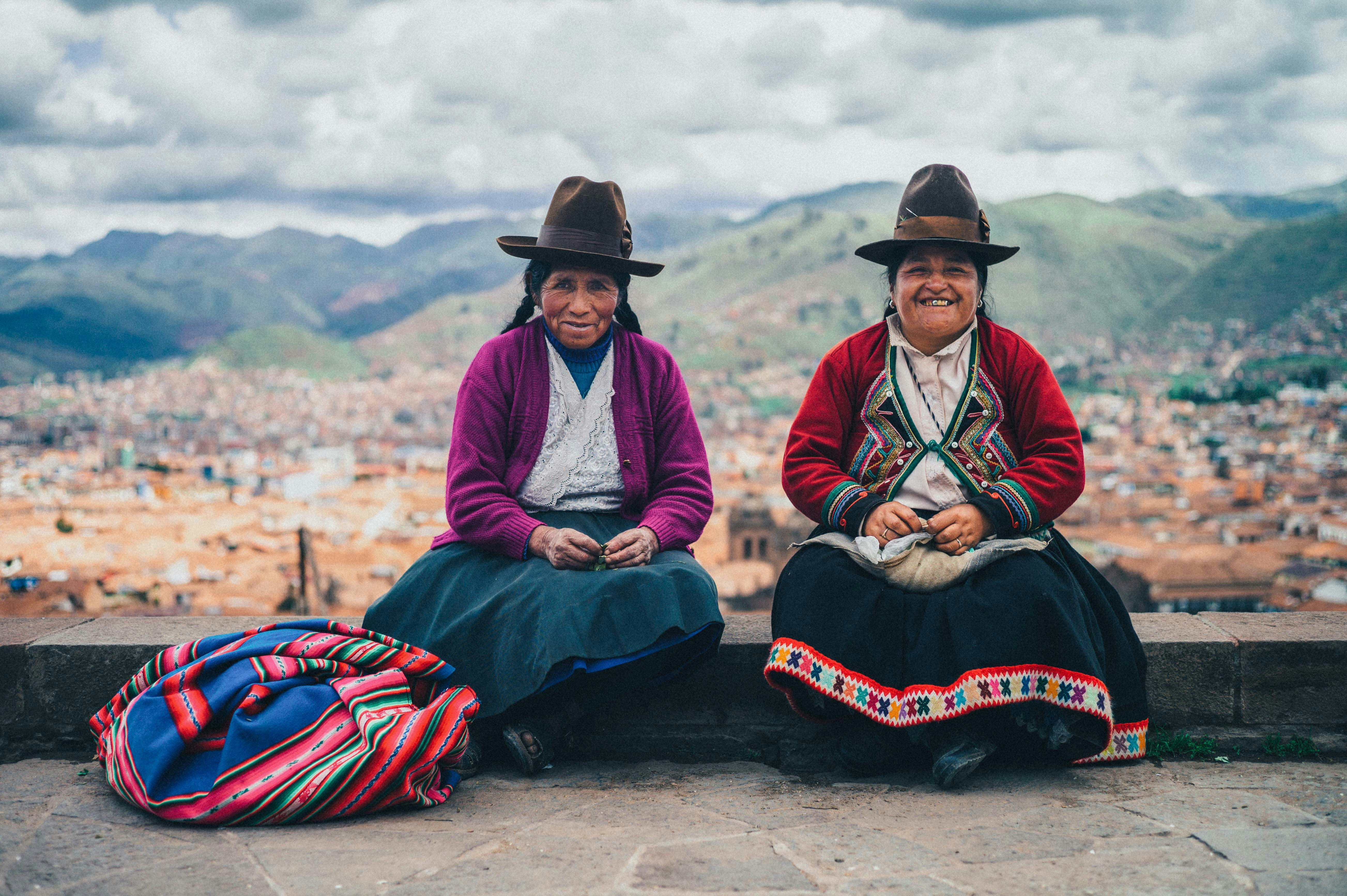 Two women in traditional Andean dress sit on a stone wall with the Andean town and hills behind; a colorful woven bag sits to the left. This is a candid street/portrait scene on a highland overlook.