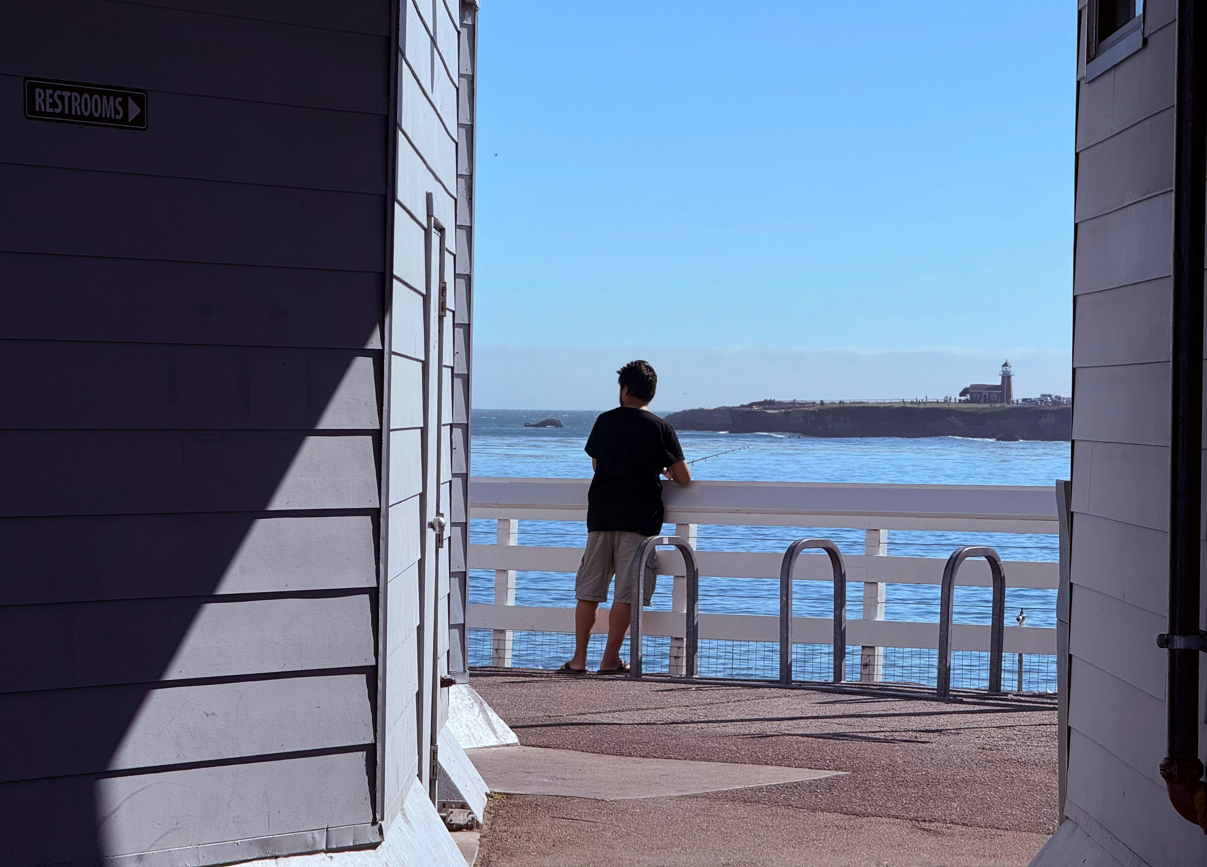 Two figures stand at the railing by the sea, framed by the entrance of a boardwalk, with a distant lighthouse on calm blue water.