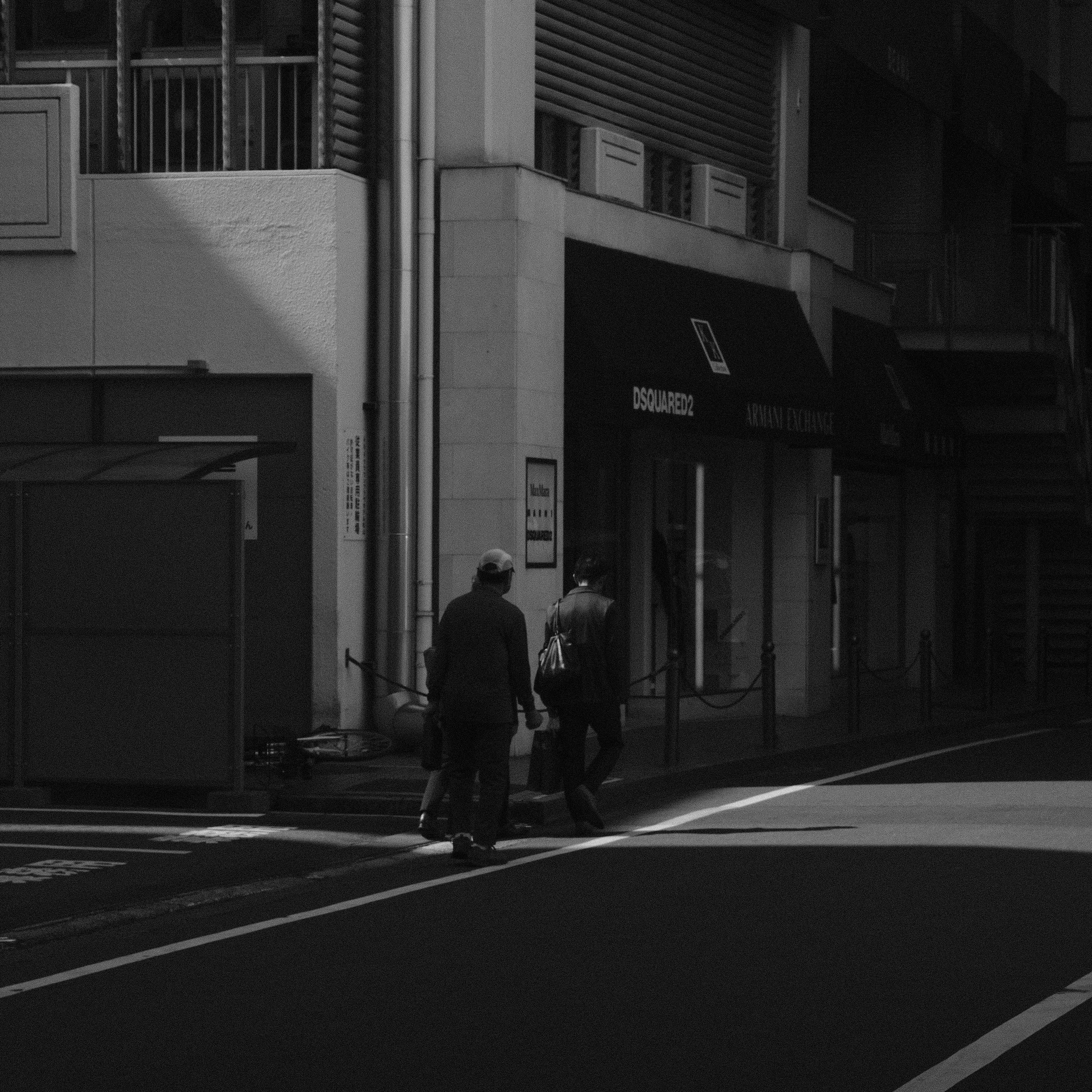 Shadowed Corner: Two pedestrians step into the sun along a quiet urban storefront