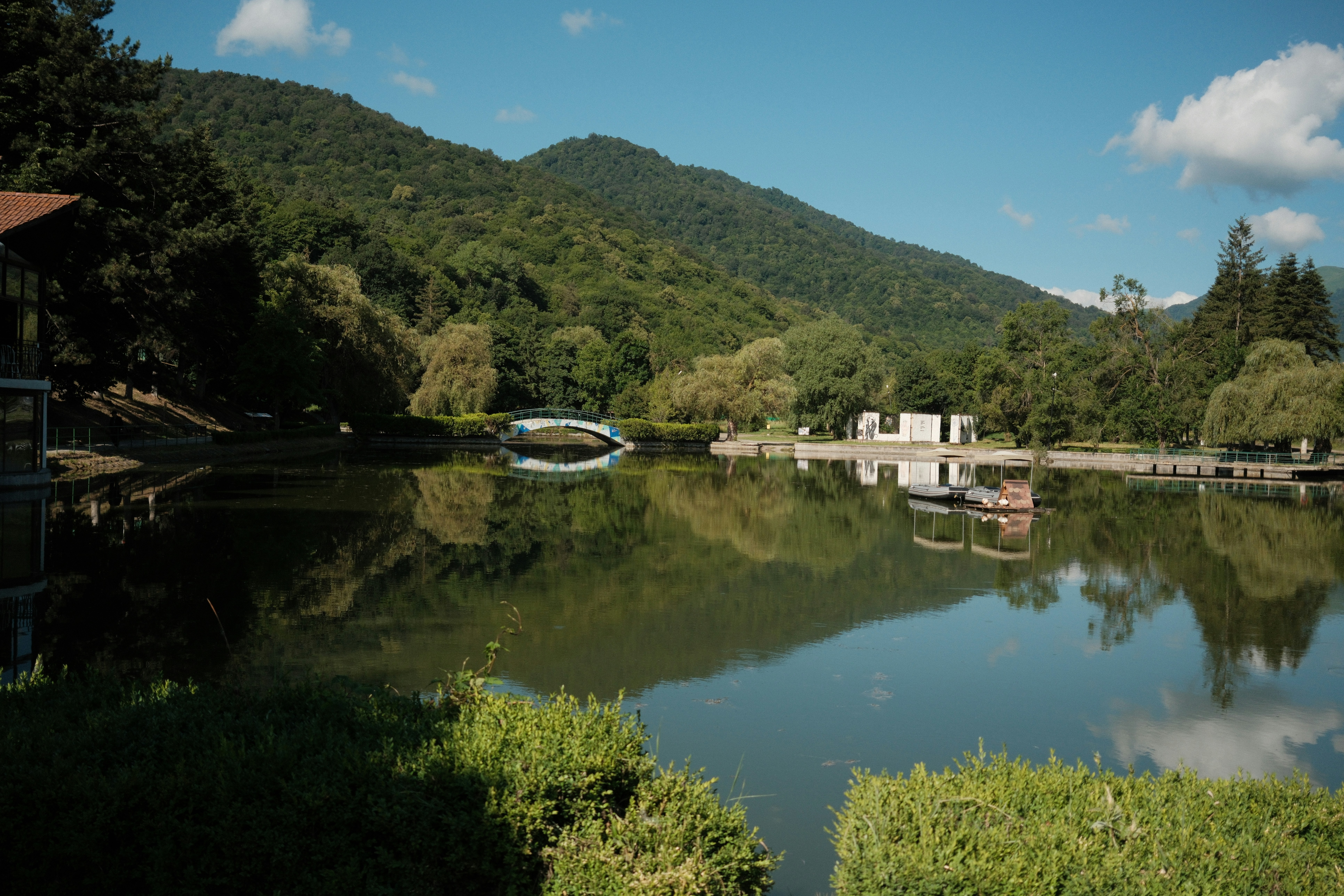 Calm lake scene with a small arched bridge and distant white structures mirrored in the water, framed by green hills and a bright blue sky.