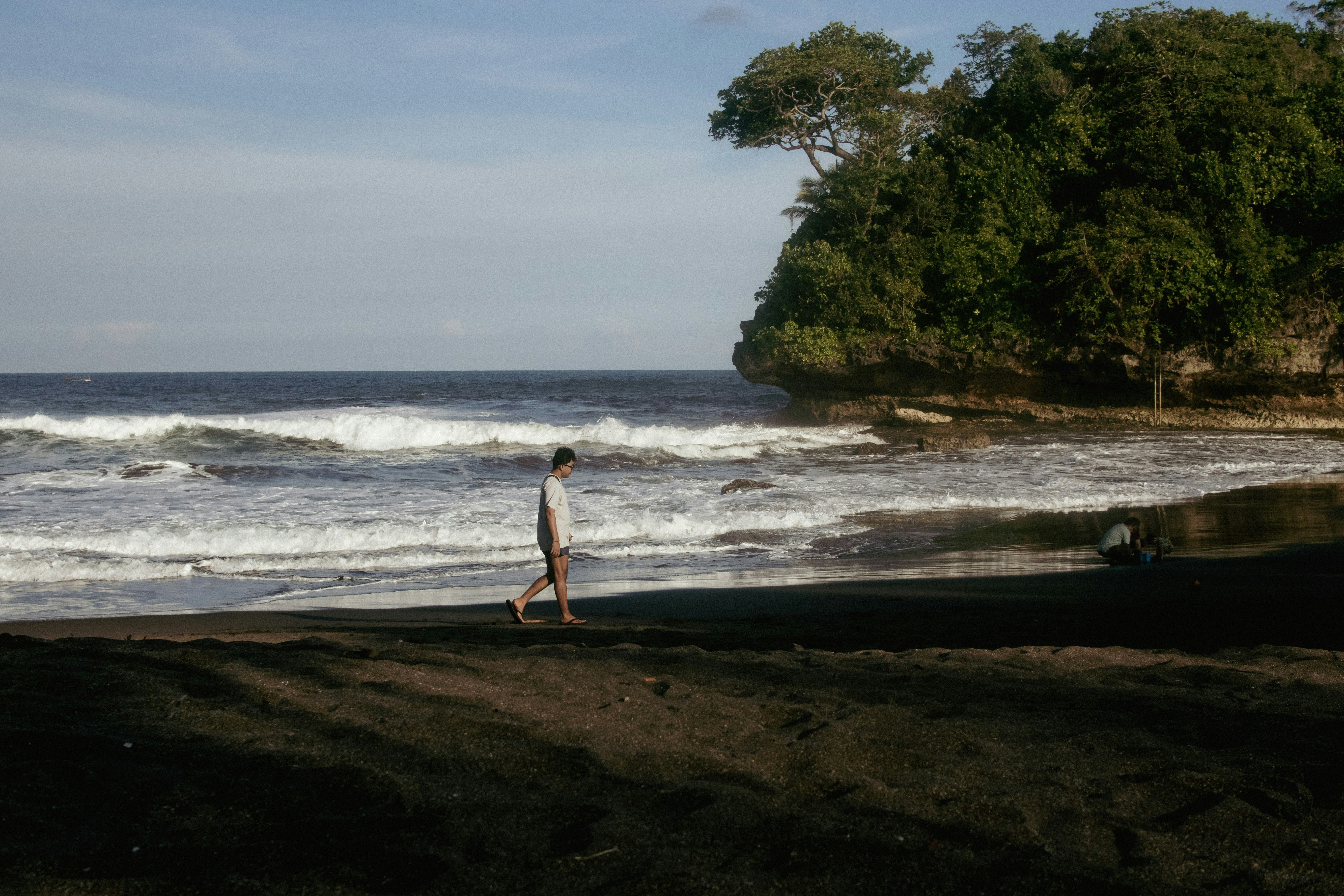 A solitary man walks along a dark sandy beach with foamy waves and a tree-covered headland in the background. This photograph captures a calm coastal scene with natural light and a hint of twilight.