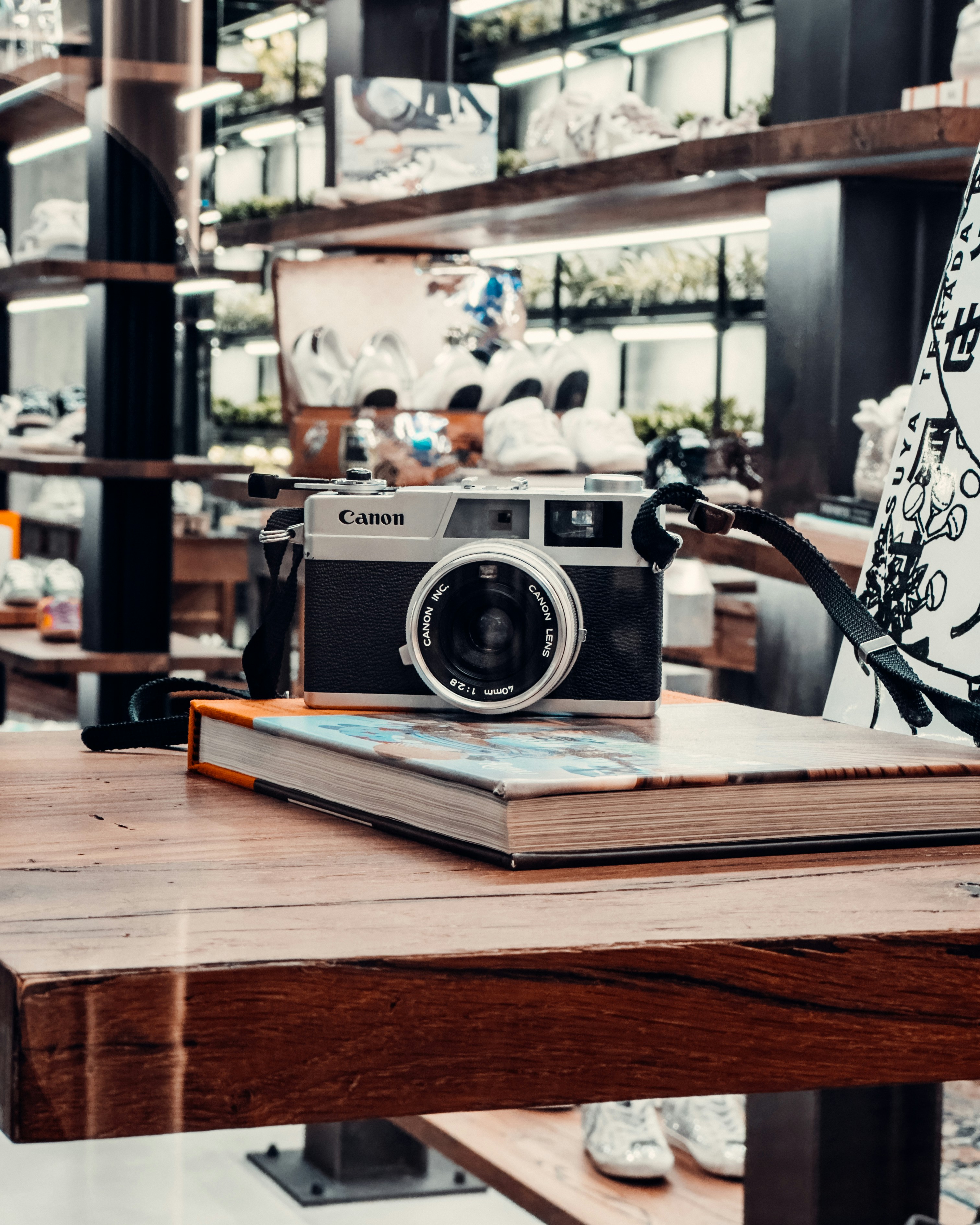 A vintage Canon camera sits atop a glossy book on a wooden table inside a store, with shelves and shoppers in the blurred background.