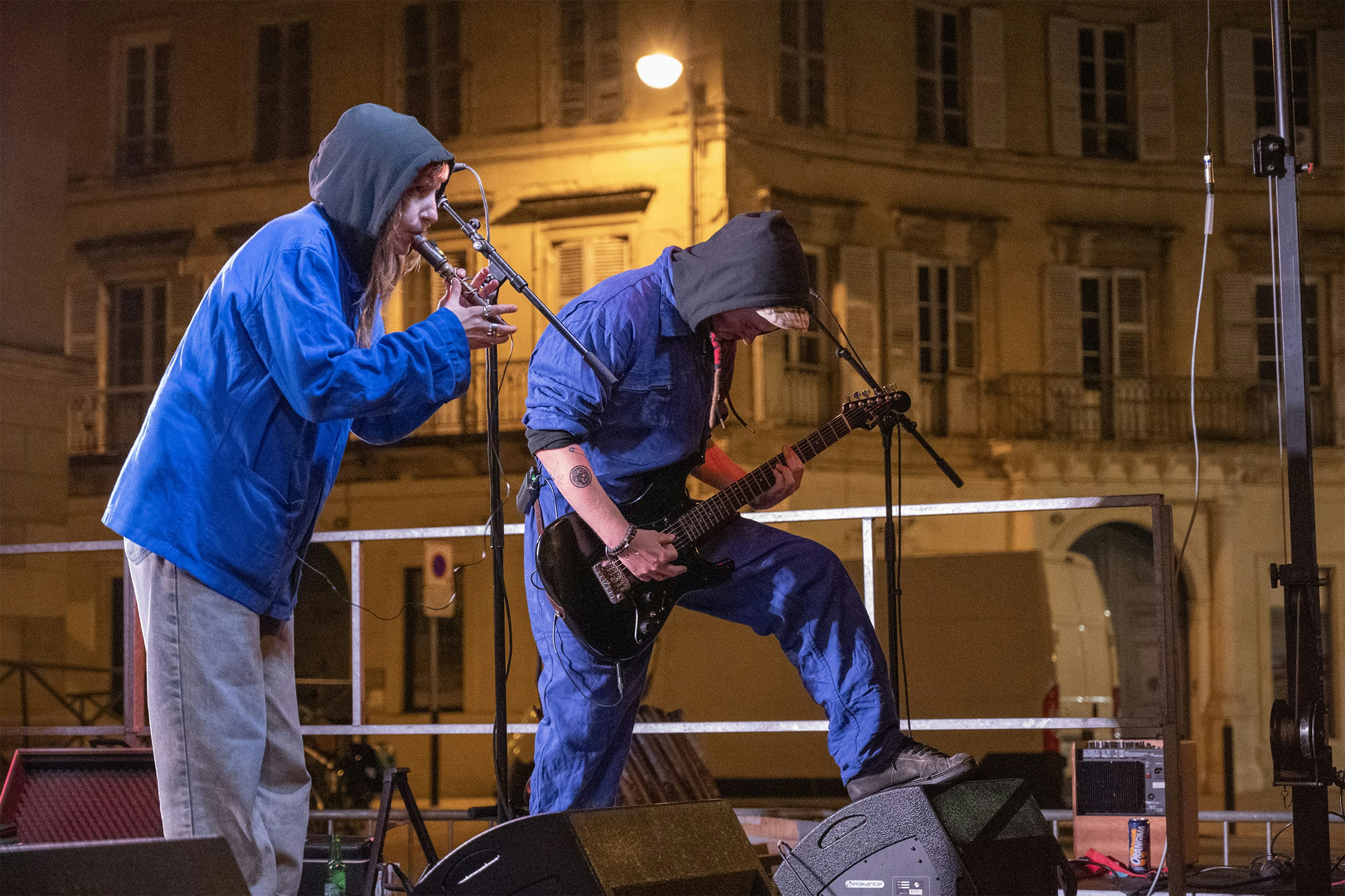 Two hooded guitarists perform on an outdoor stage at night, blue hoodies and jackets contrasted against warm urban lighting; one guitarist leans into a riff while the other engages with a mic, cables and amps hinting at a live street show.