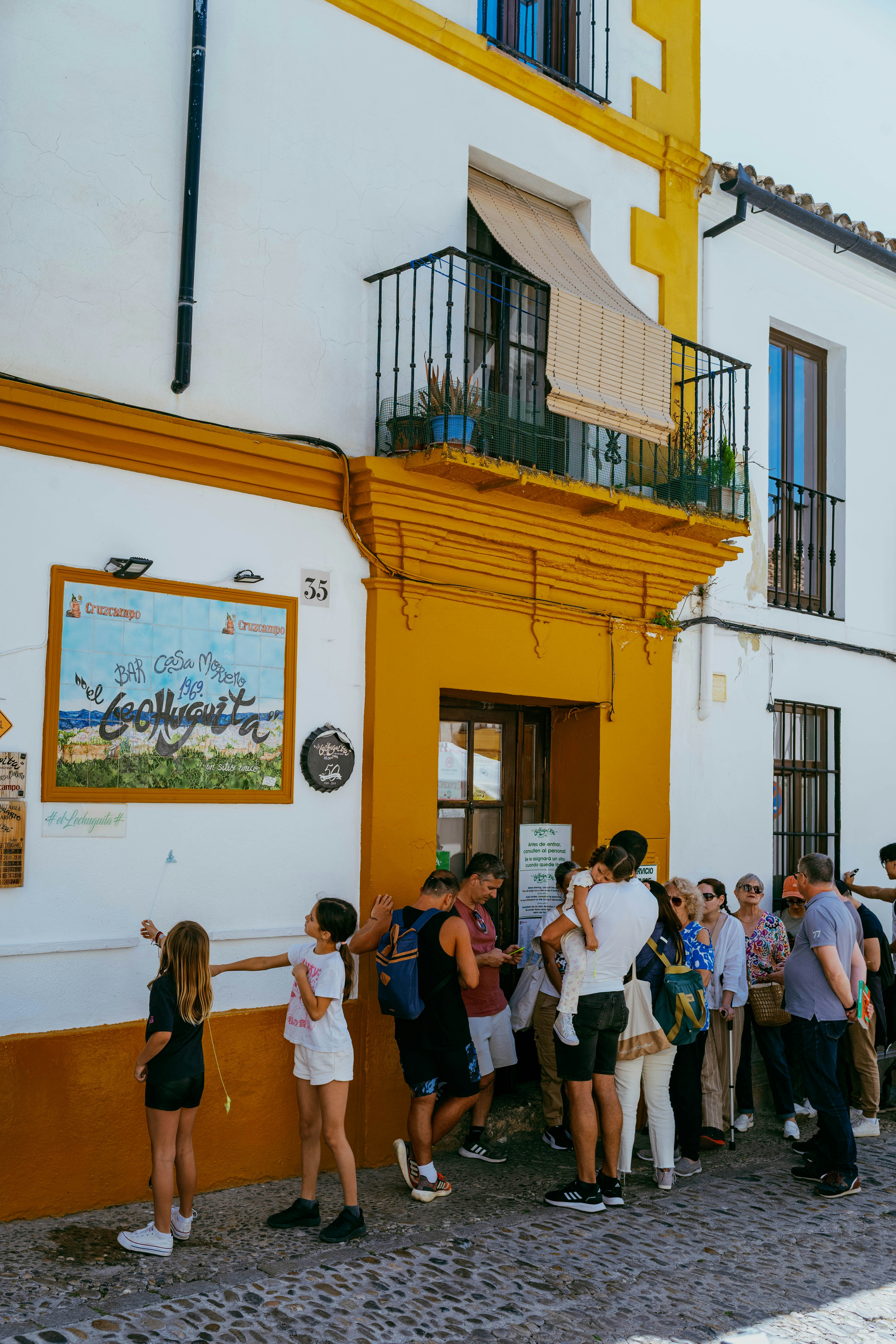 Queue at a sunlit Andalusian doorway by a yellow-walled cafe