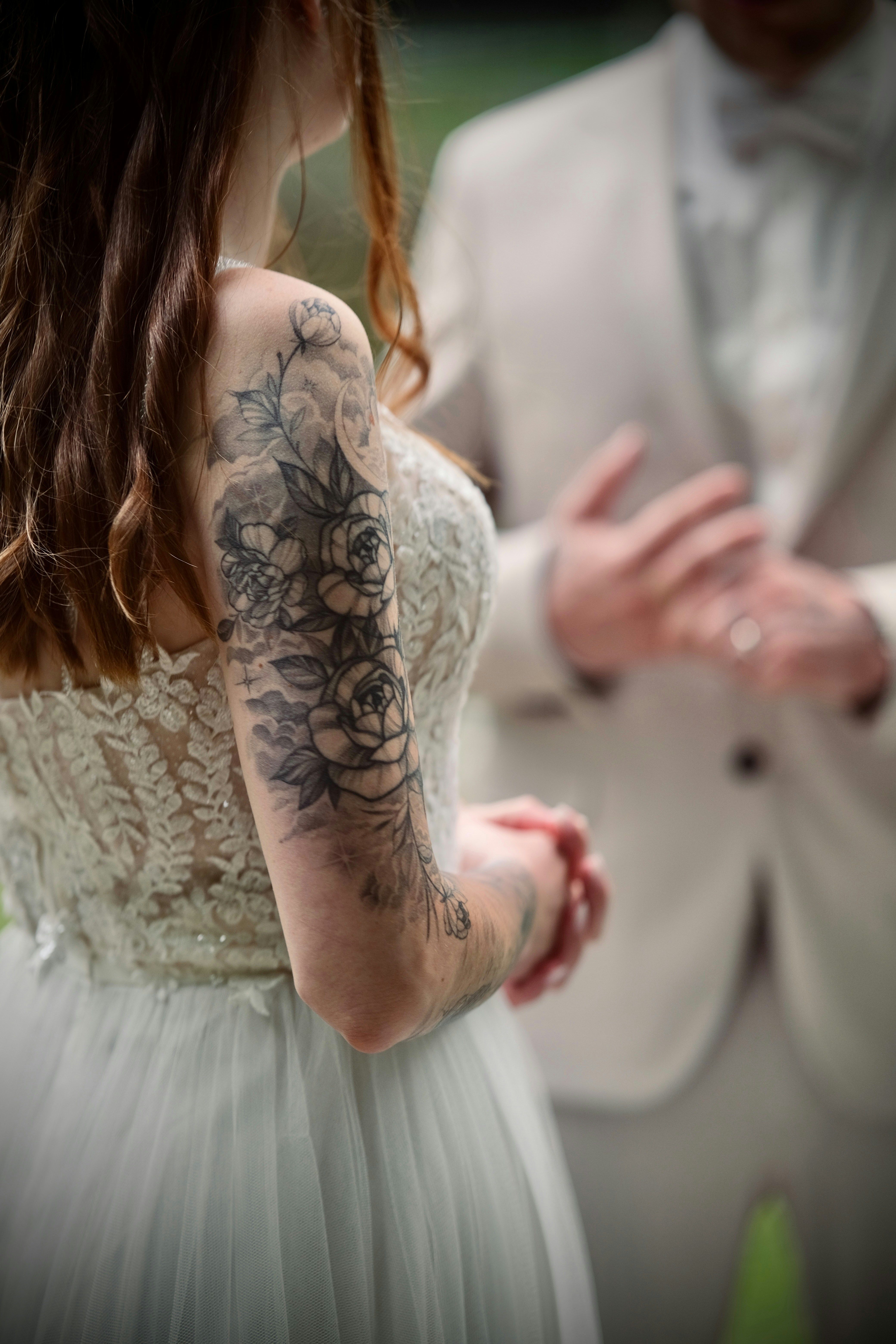 Bride with Lace Dress, Tattooed Arm, and Blurred Groom in soft daylight