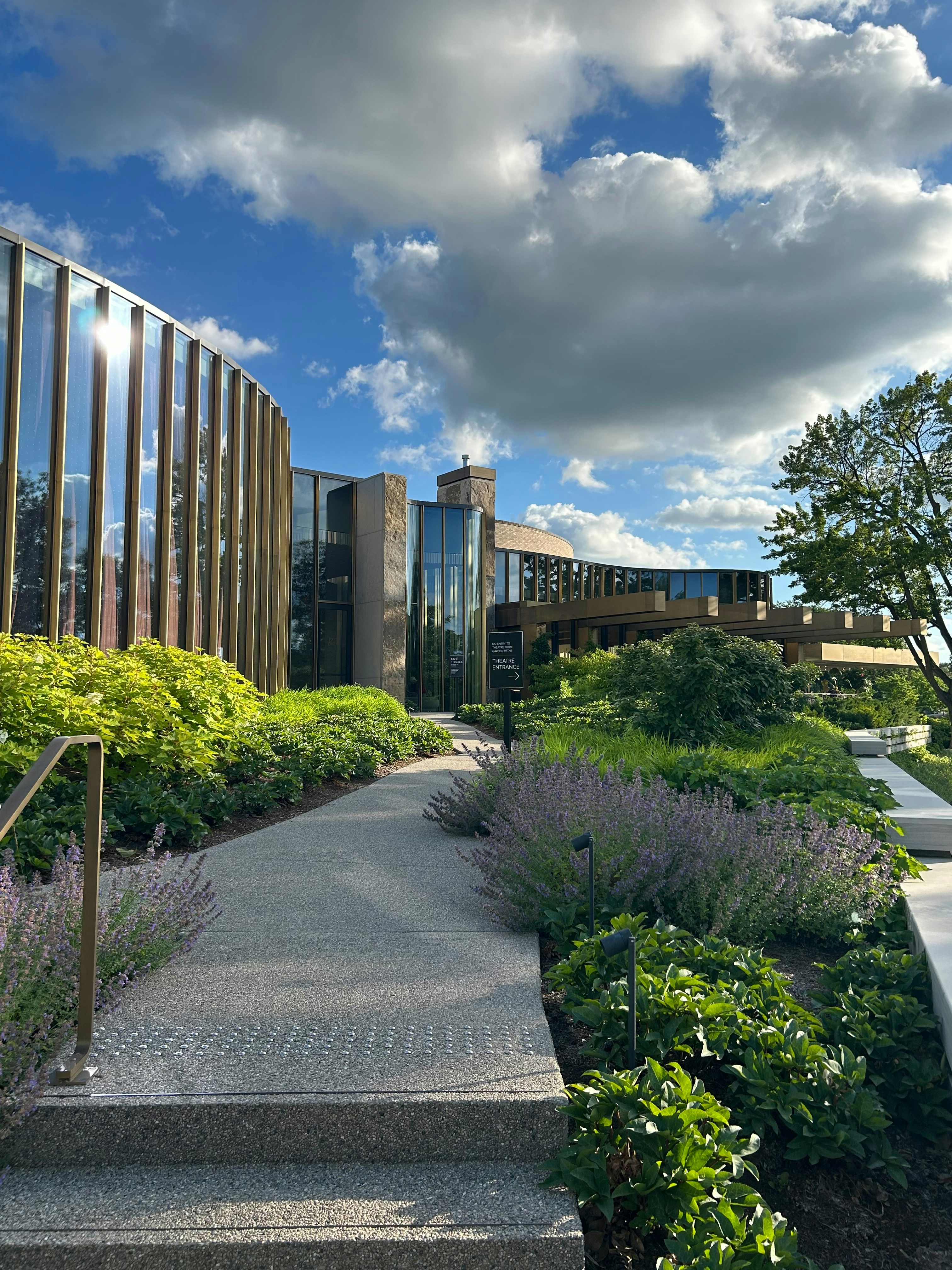 Sculpted walkway lined with lush greenery leads to a curved glass-fronted building under a bright sky.