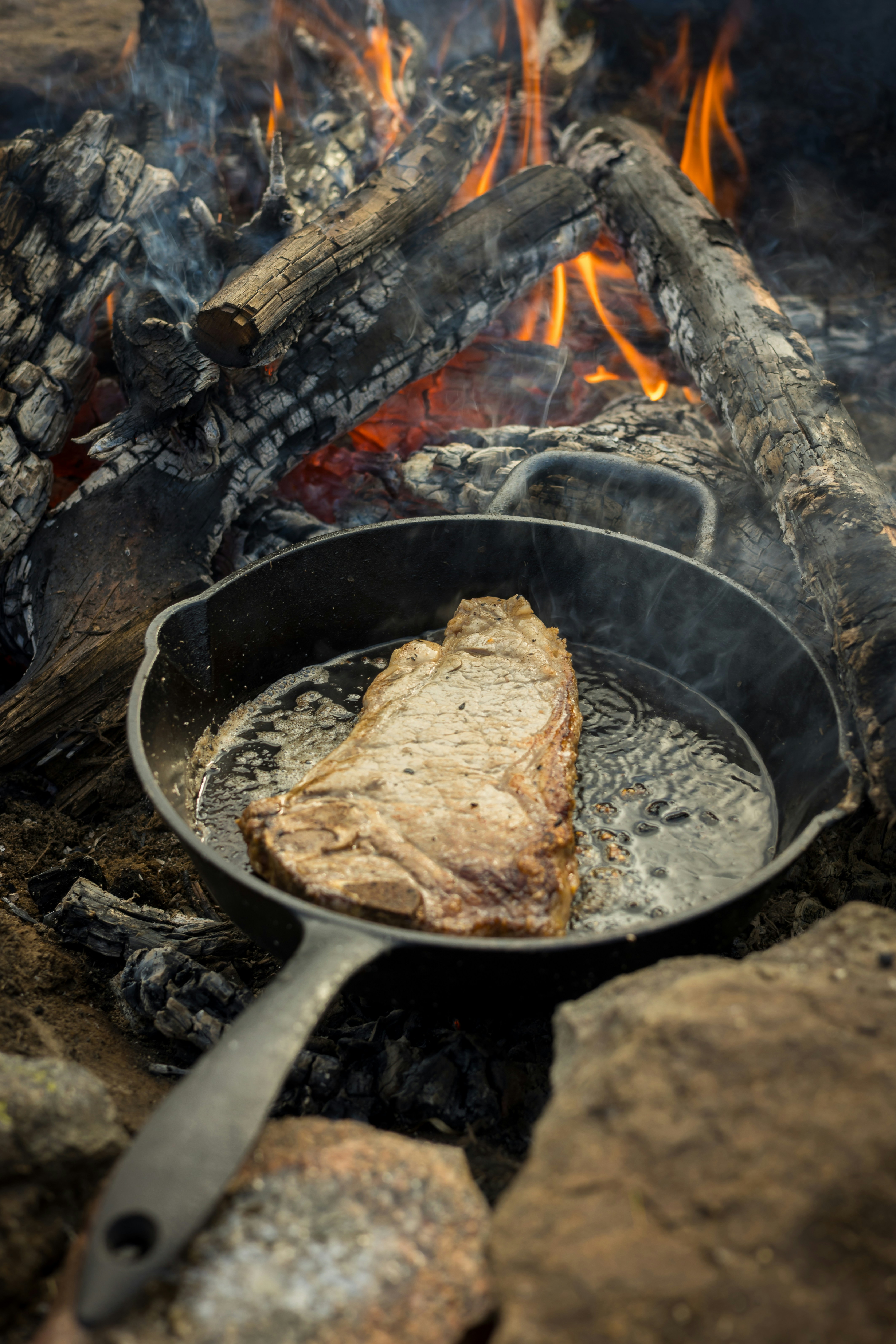 A cast iron pan with a chop resting in hot butter sits over glowing embers, surrounded by charred wood and flames, suggesting a rustic outdoor cooking scene.