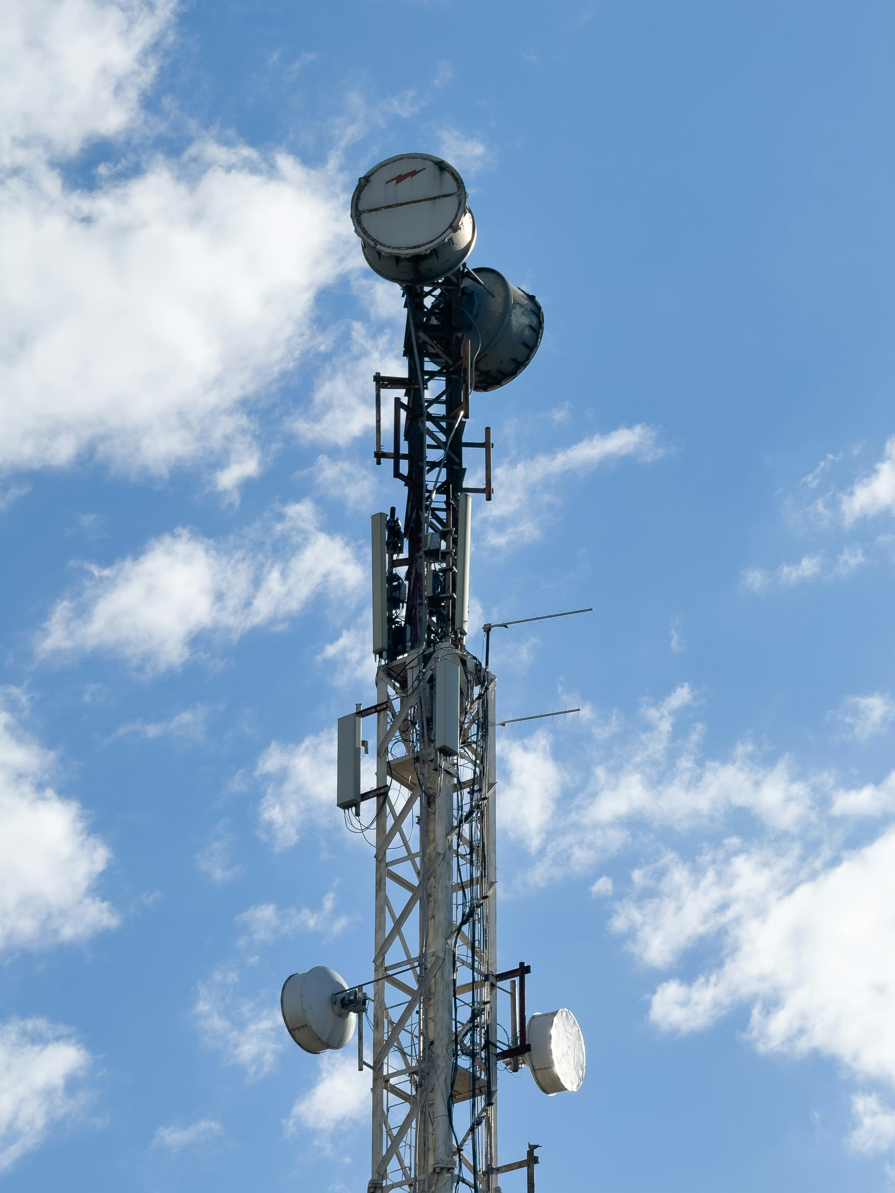 Sleek cellular tower rising into a clear blue sky with scattered clouds, featuring antennas and dish panels. This image captures the structural details and hardware of the mast.