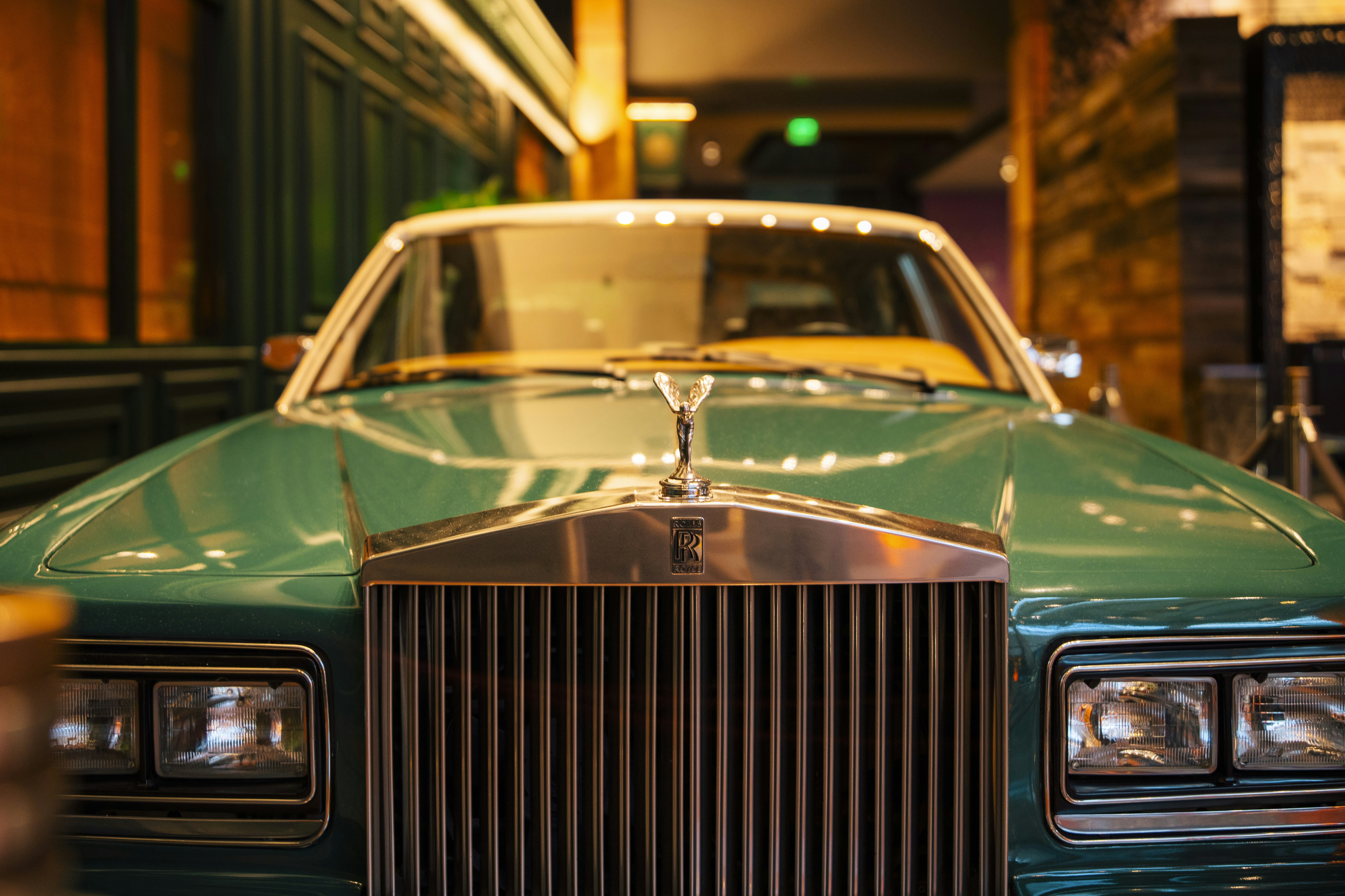 A vintage teal Rolls-Royce grille dominates a dim, warm-lit interior; the emblematic hood ornament sits center as the car faces forward, inviting a closer look at its chrome details and rich color.