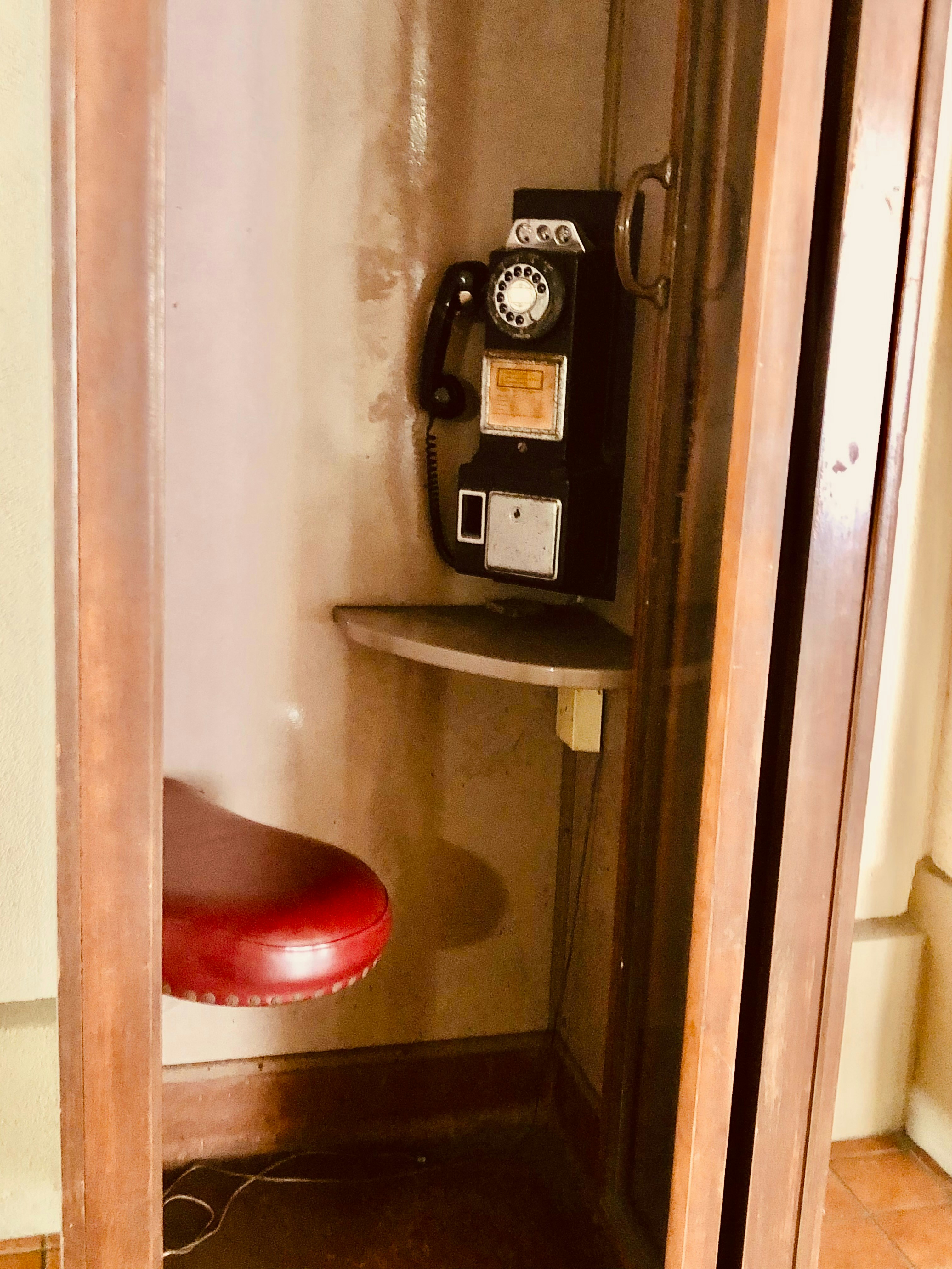 A vintage wall-mounted payphone inside a narrow wooden booth, with a red cushioned seat and warm lighting; a solitary retro device anchored to a beige wall, suggesting an old, quiet corridor.