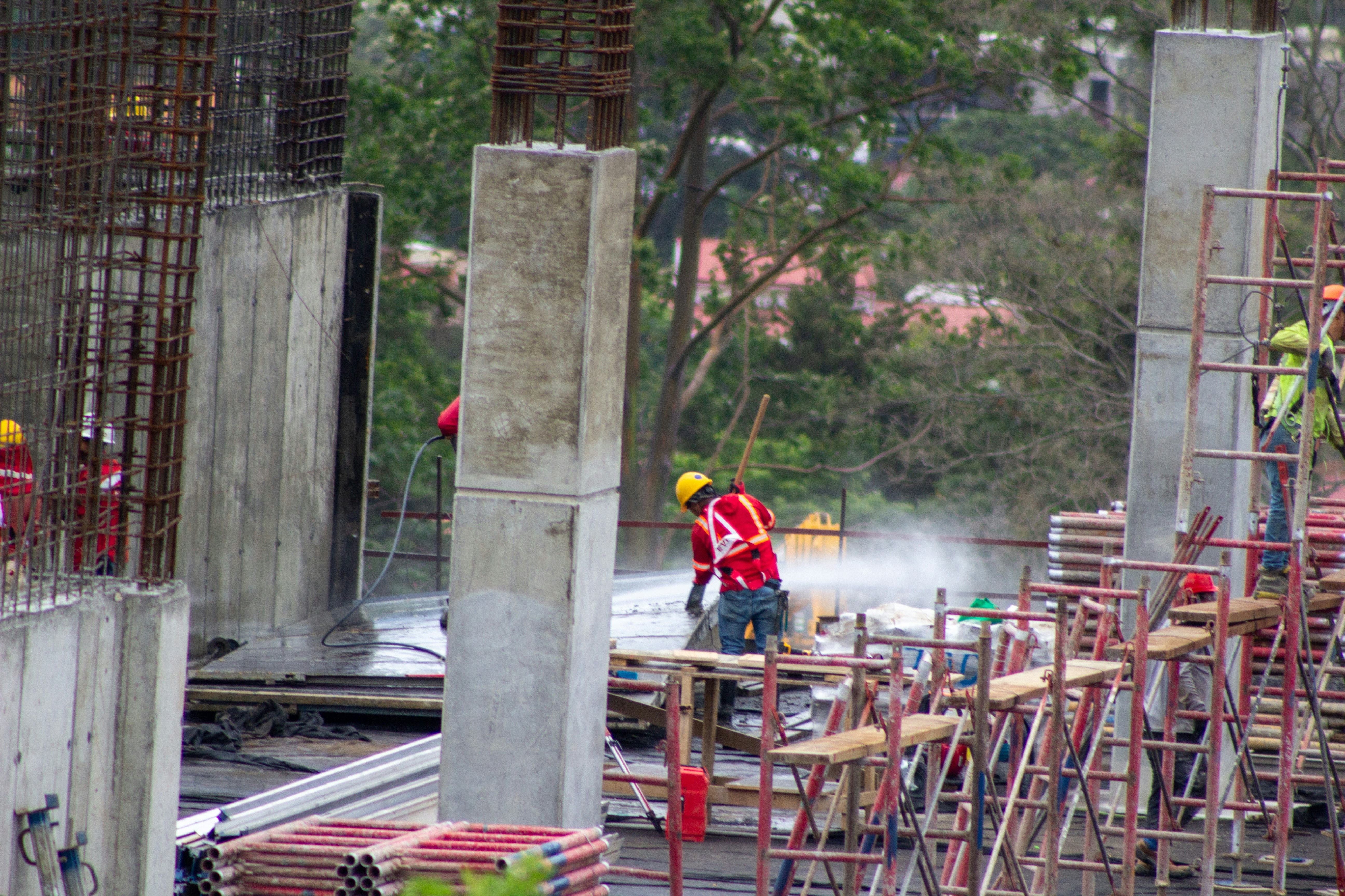 Construction workers in yellow helmets working amidst concrete pillars and scaffolding on a partially built structure; a misty, busy site setting with reinforcements and materials visible in the foreground.