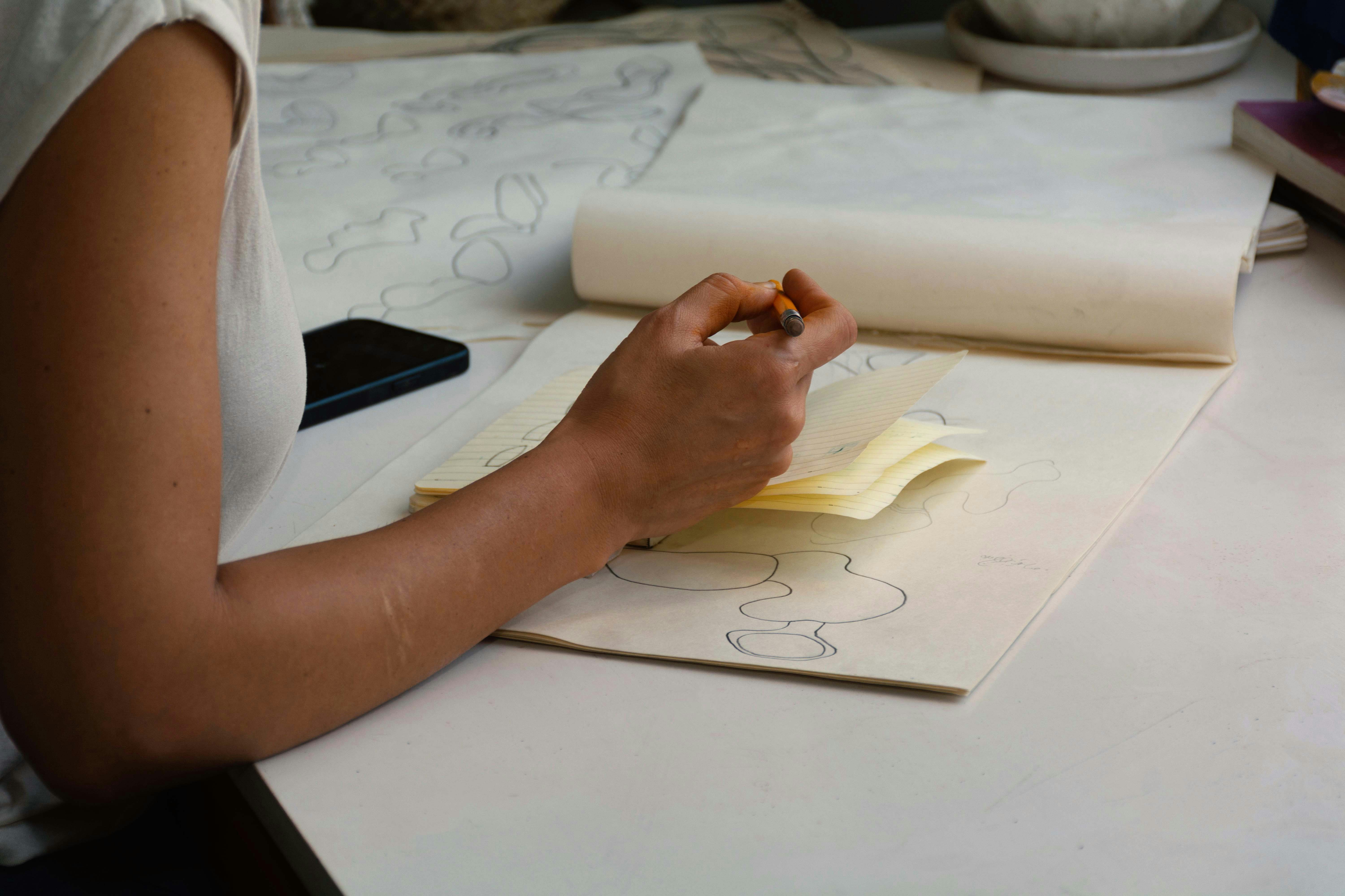 A close-up of a person sketching into a notebook on a white desk, with a smartphone nearby and a rolled sheet in the background. The scene emphasizes the act of drawing and the workspace.