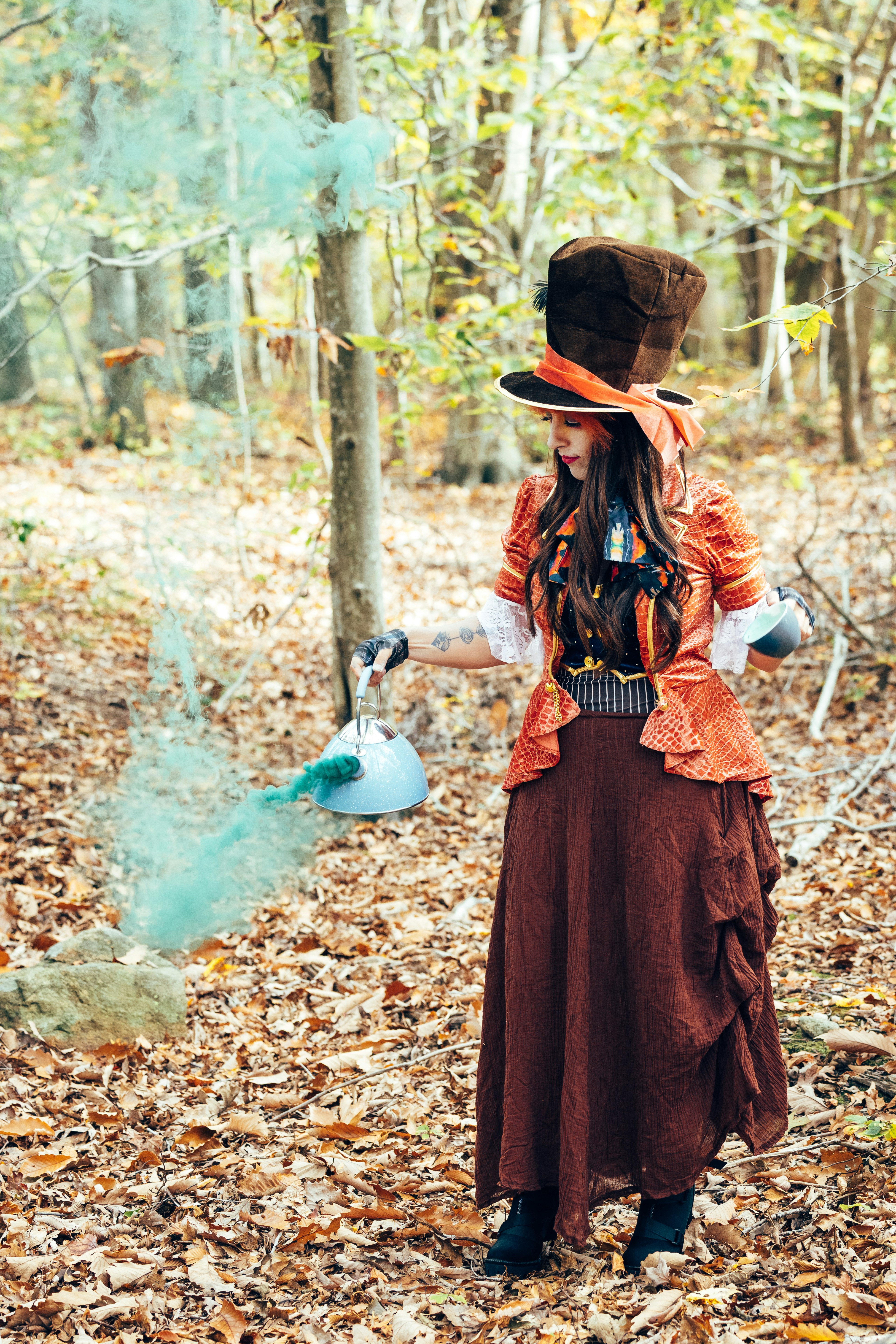 Autumn scene of a person in a vintage costume releasing teal smoke in a leaf-strewn forest; the moment centers on the figure’s upper body and imaginary haze, highlighting color and texture. Logo-like branding not present.