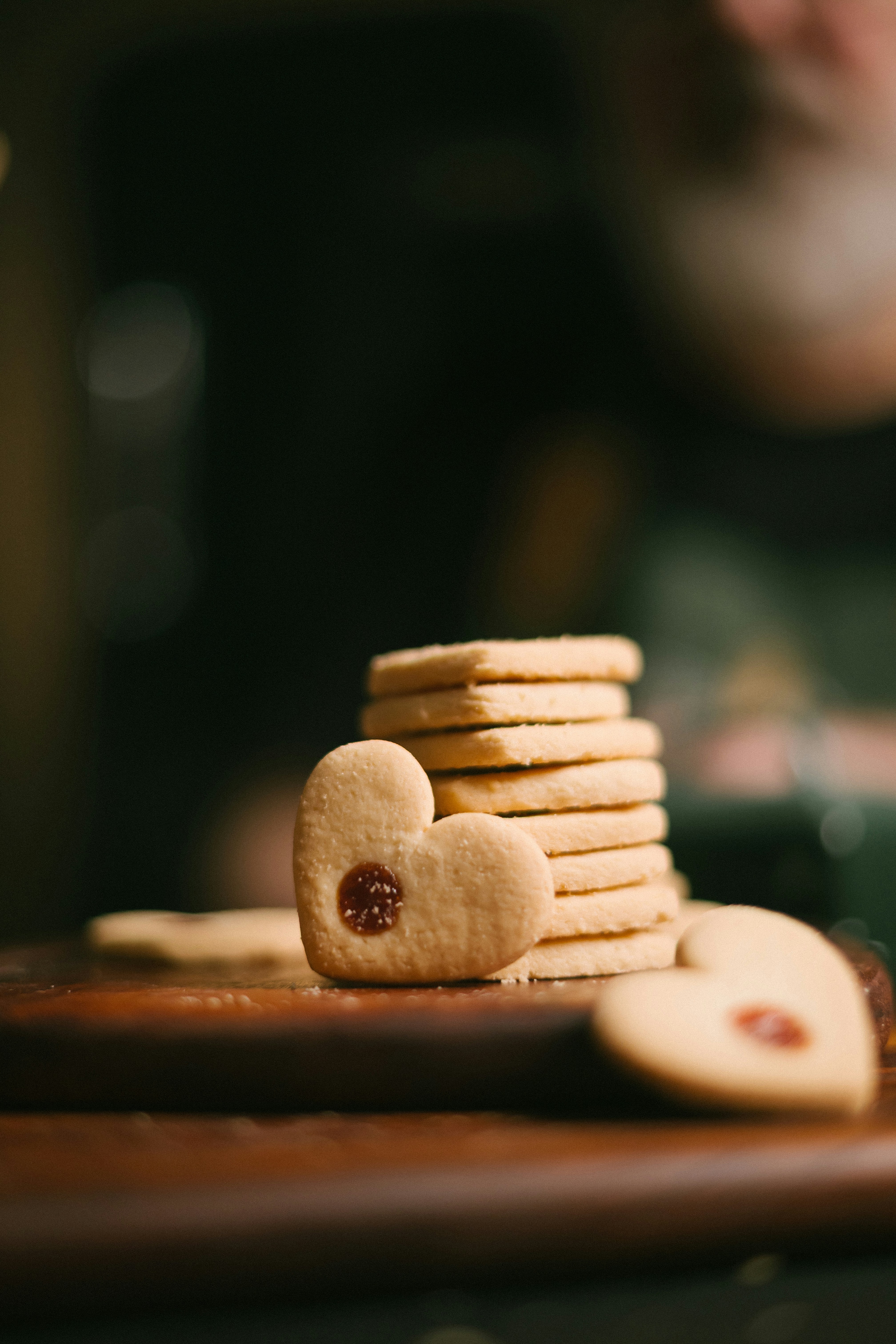 A stack of heart-shaped cookies with jam centers sits on a wooden board, foregrounded with one cookie leaning against the stack; soft, warm lighting highlights the pastry while a blurred background suggests a kitchen scene.