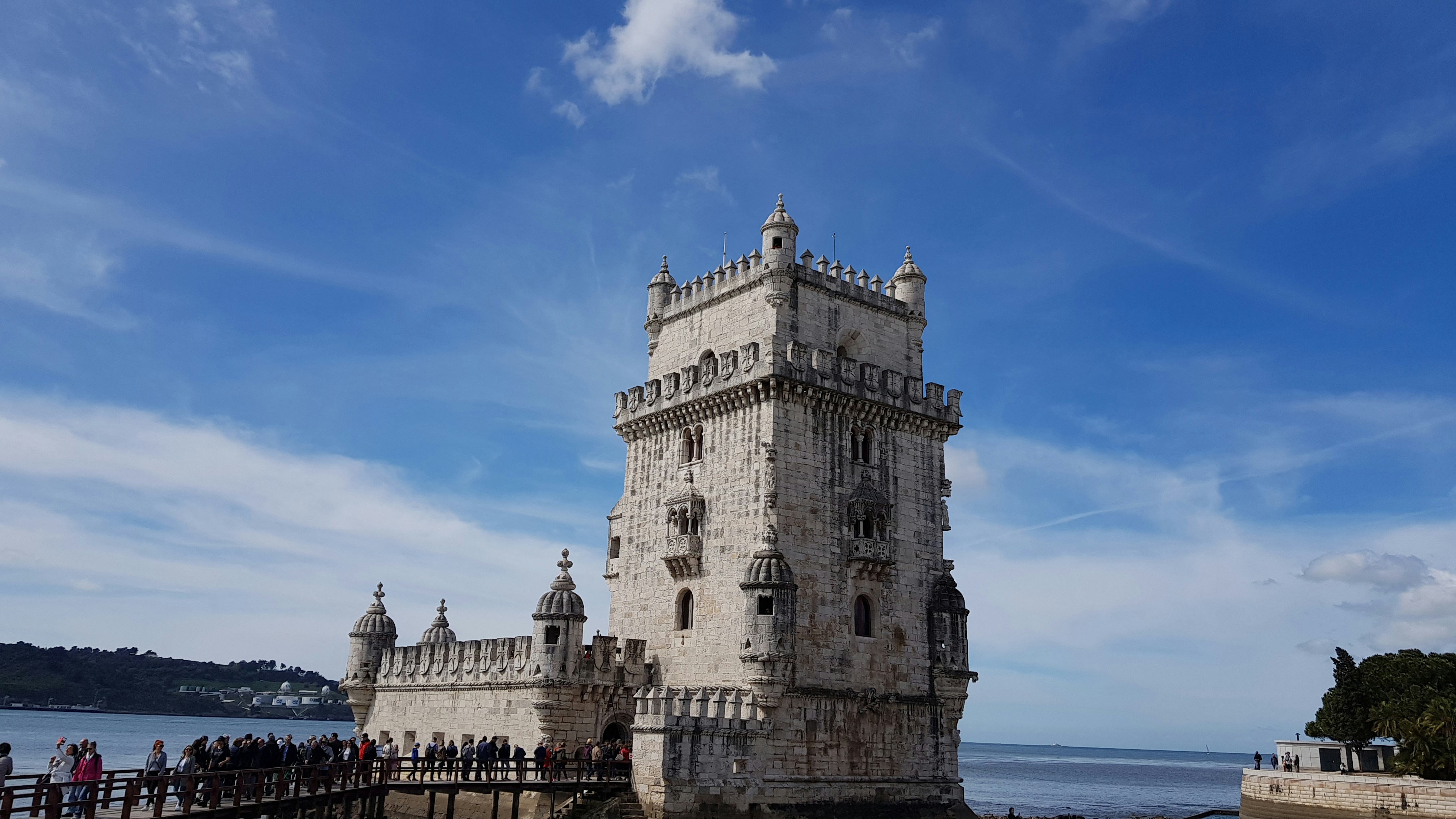 Medieval fortress towers above a pebble promenade along a calm Atlantic shoreline, with visitors near the base and a bright blue sky. The scene highlights maritime architecture and coastal tourism.