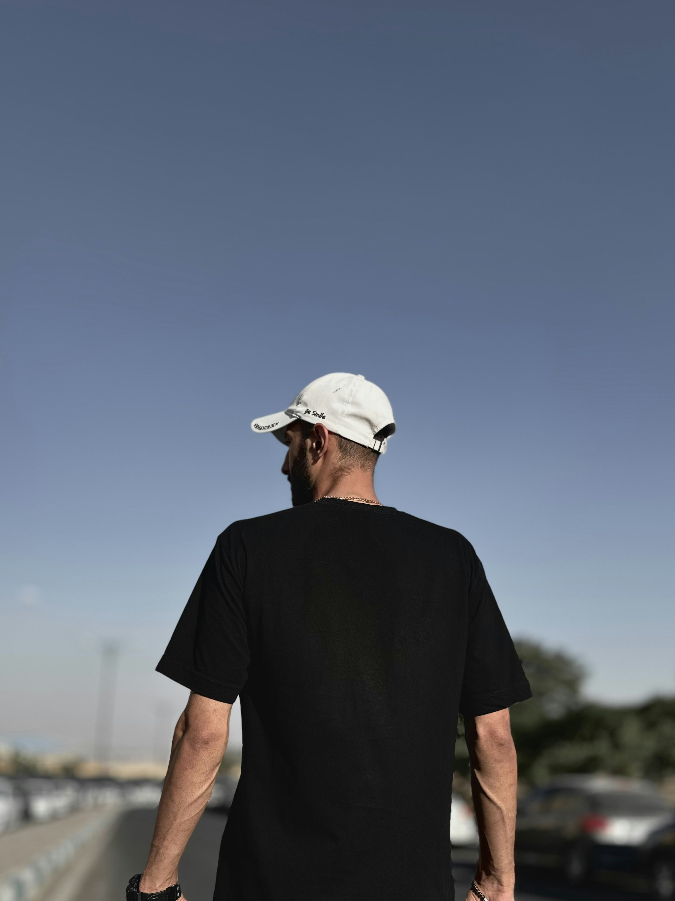 A man in a black shirt and white cap stands with his back to the camera, facing a clear blue sky and a distant roadside scene.
