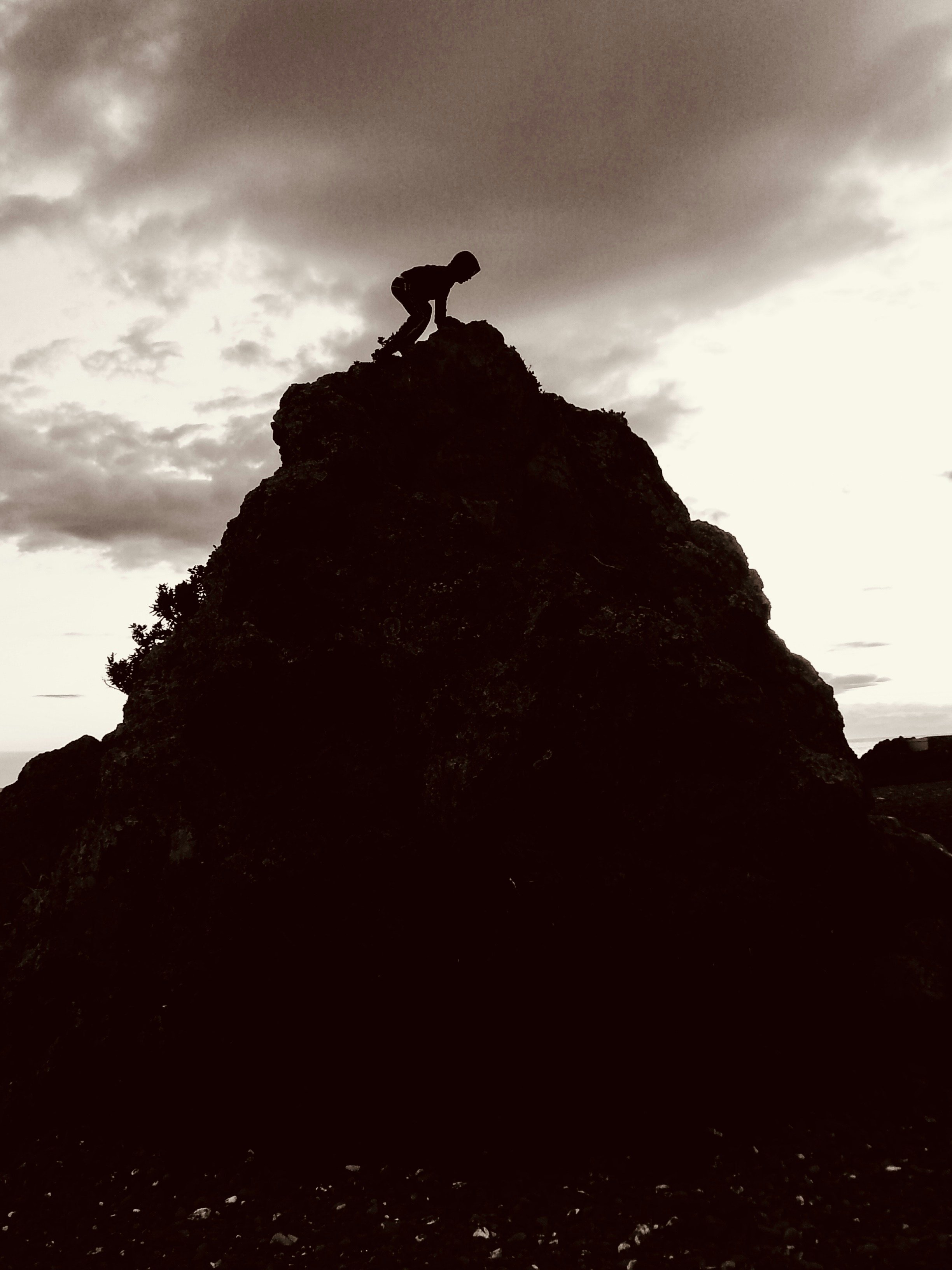 Dark silhouette of a climber atop a jagged rock formation against a cloudy sky; the image emphasizes scale and solitude with a high-contrast, monochromatic tone. (Black-and-white silhouette, dramatic landscape art).